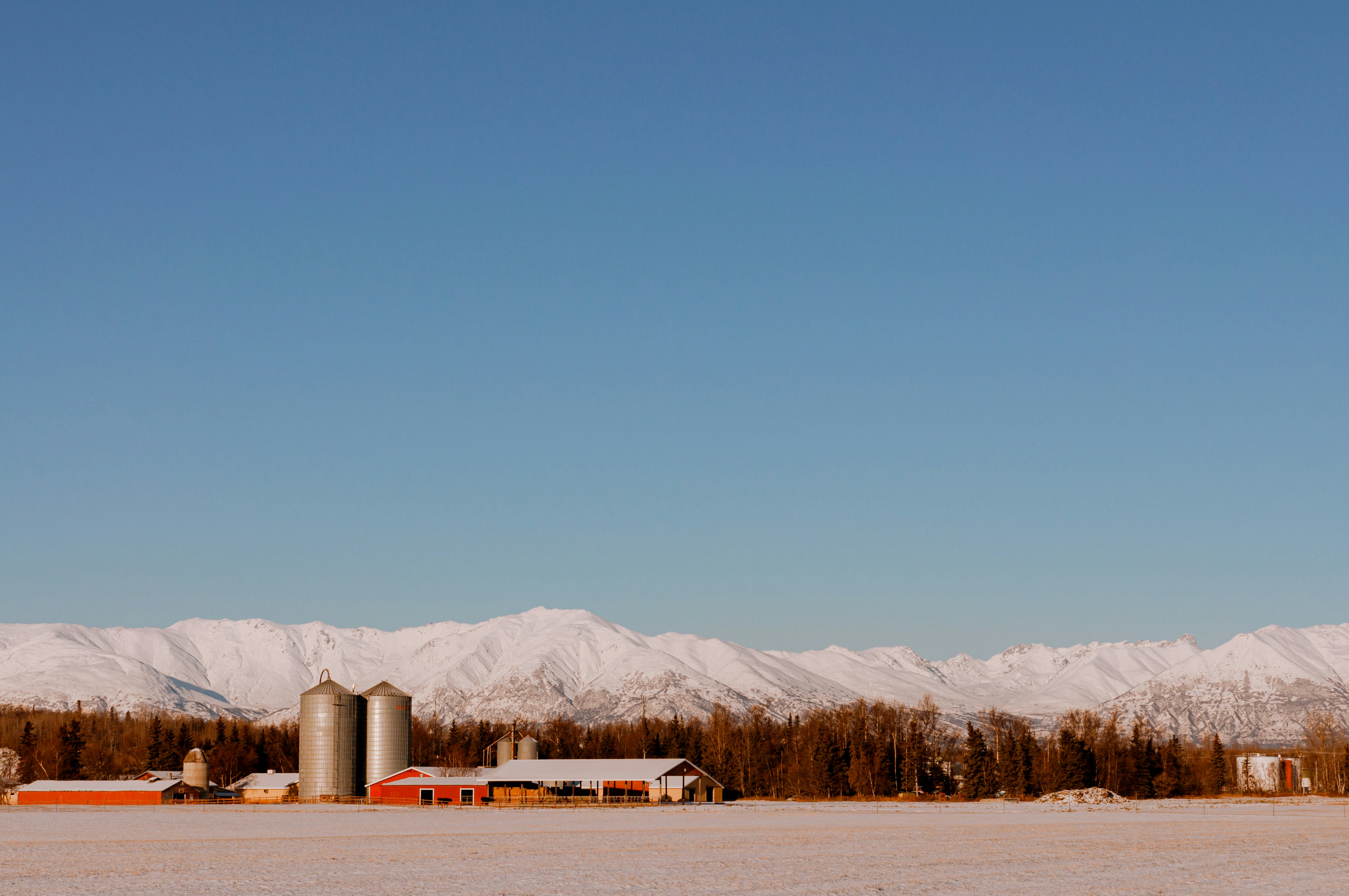 Snow-covered farm with silos and barns set against a backdrop of majestic mountains under a clear blue sky.