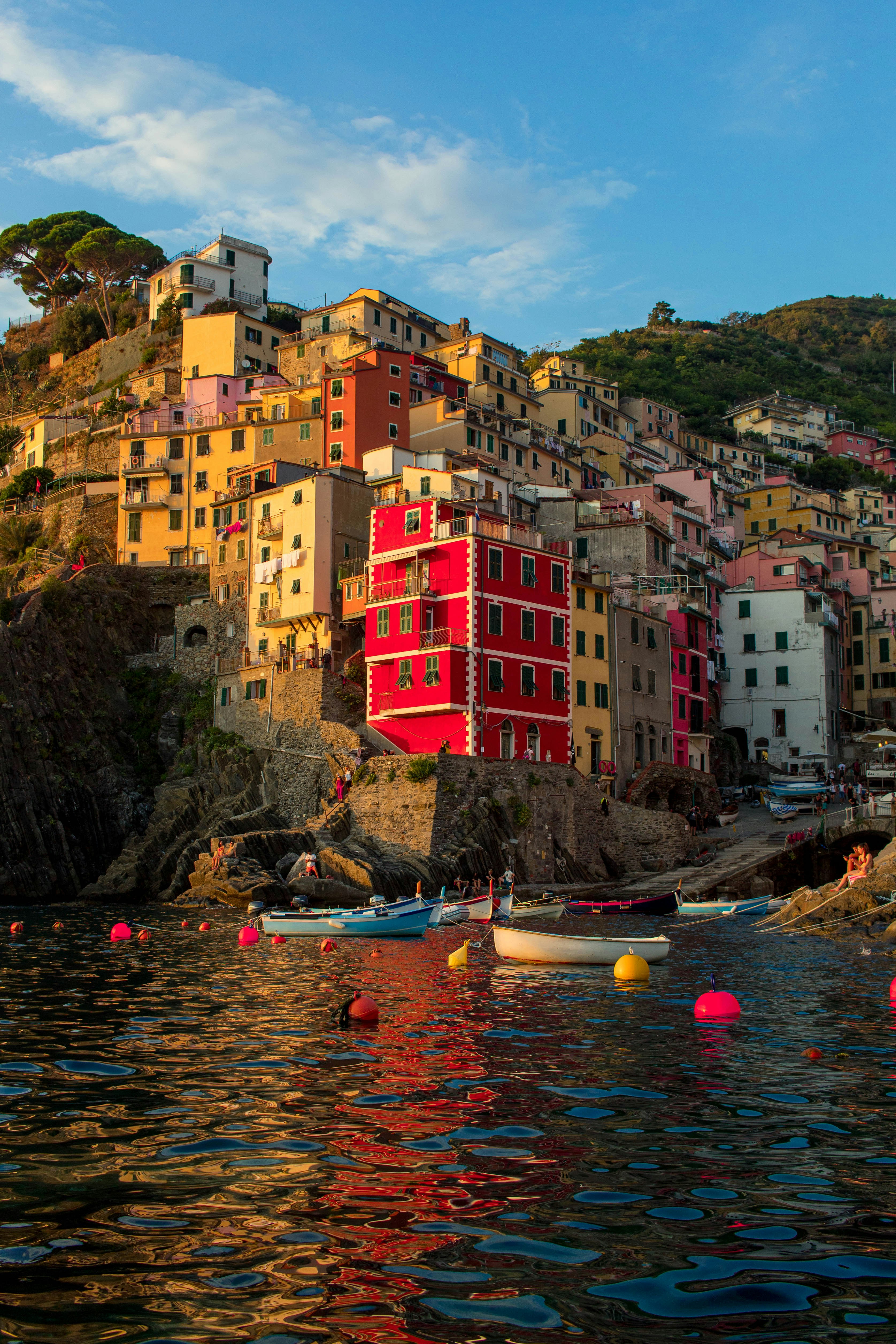 Colorful houses stacked on a hillside above a tranquil harbor, reflecting golden hues in the water. Brightly colored buoys float nearby.