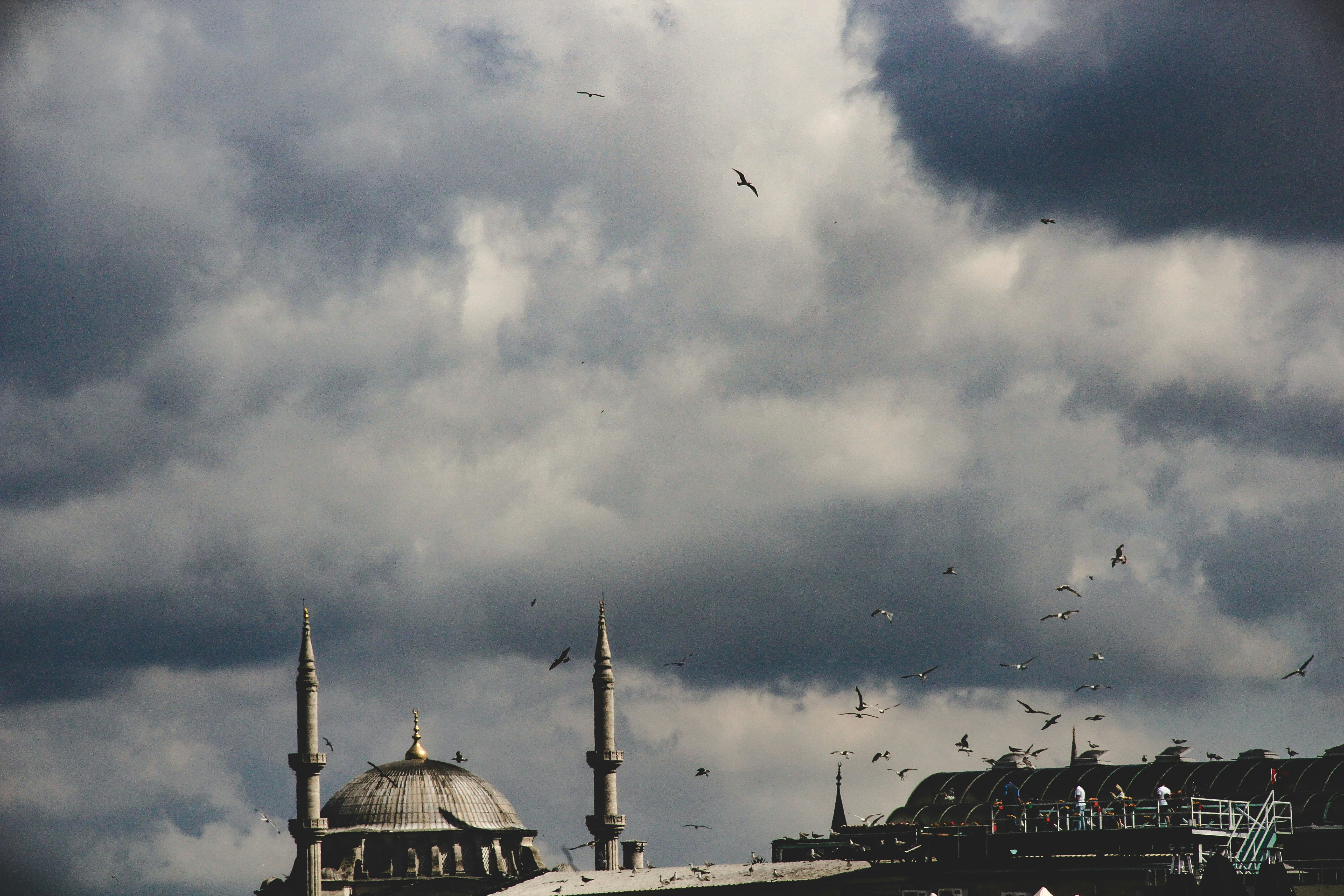Dramatic clouds loom over a historic mosque and cityscape, with birds soaring beneath the turbulent sky.