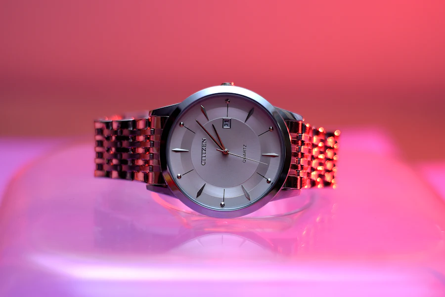 Silver Citizen wristwatch centered on a glossy pink surface with a soft magenta background and reflections below. The metal bracelet and clean analog dial are highlighted by studio lighting.