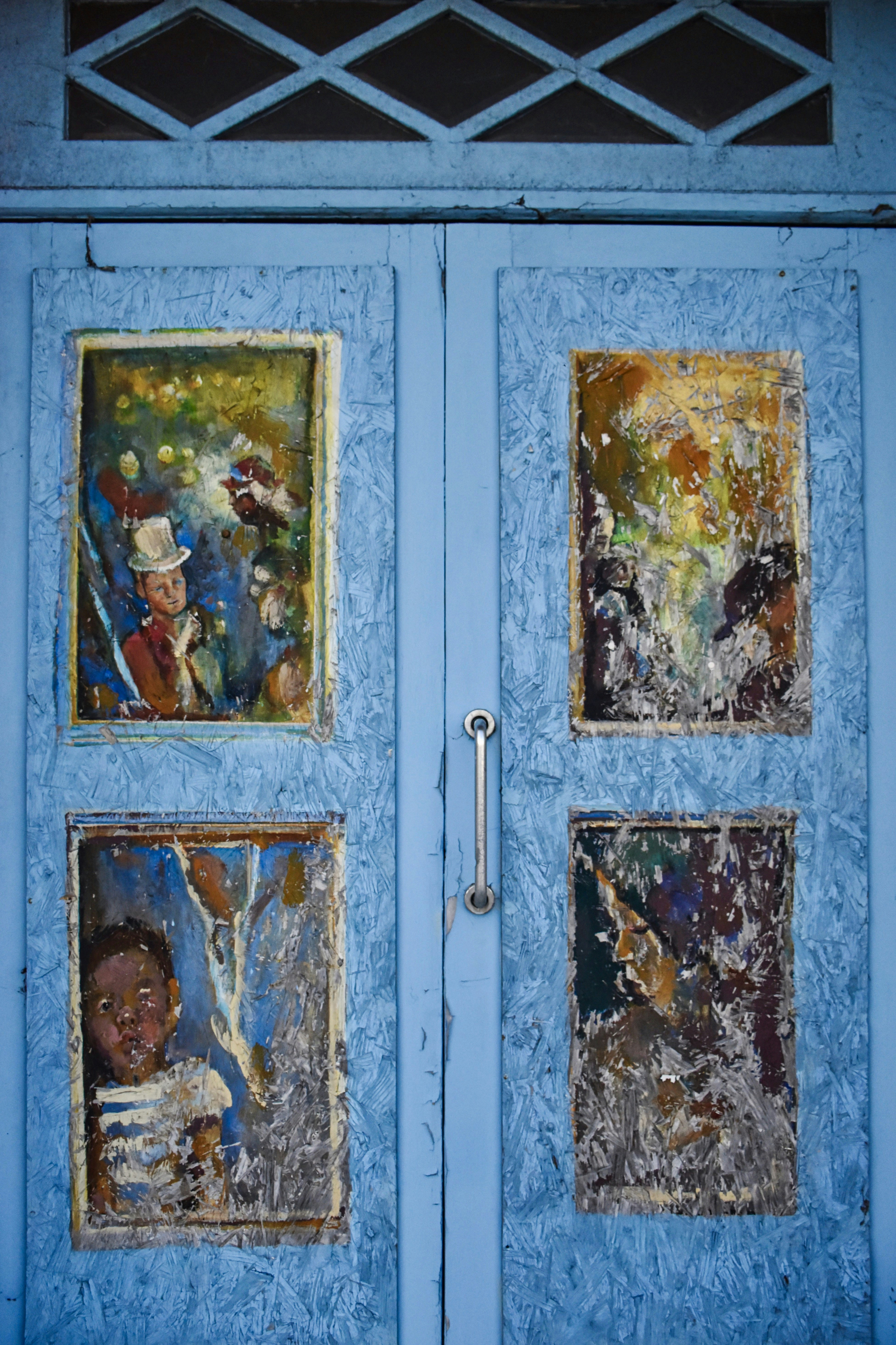 Artistic door adorned with four framed paintings, featuring vibrant colors and diverse subjects, set against a textured blue background.
