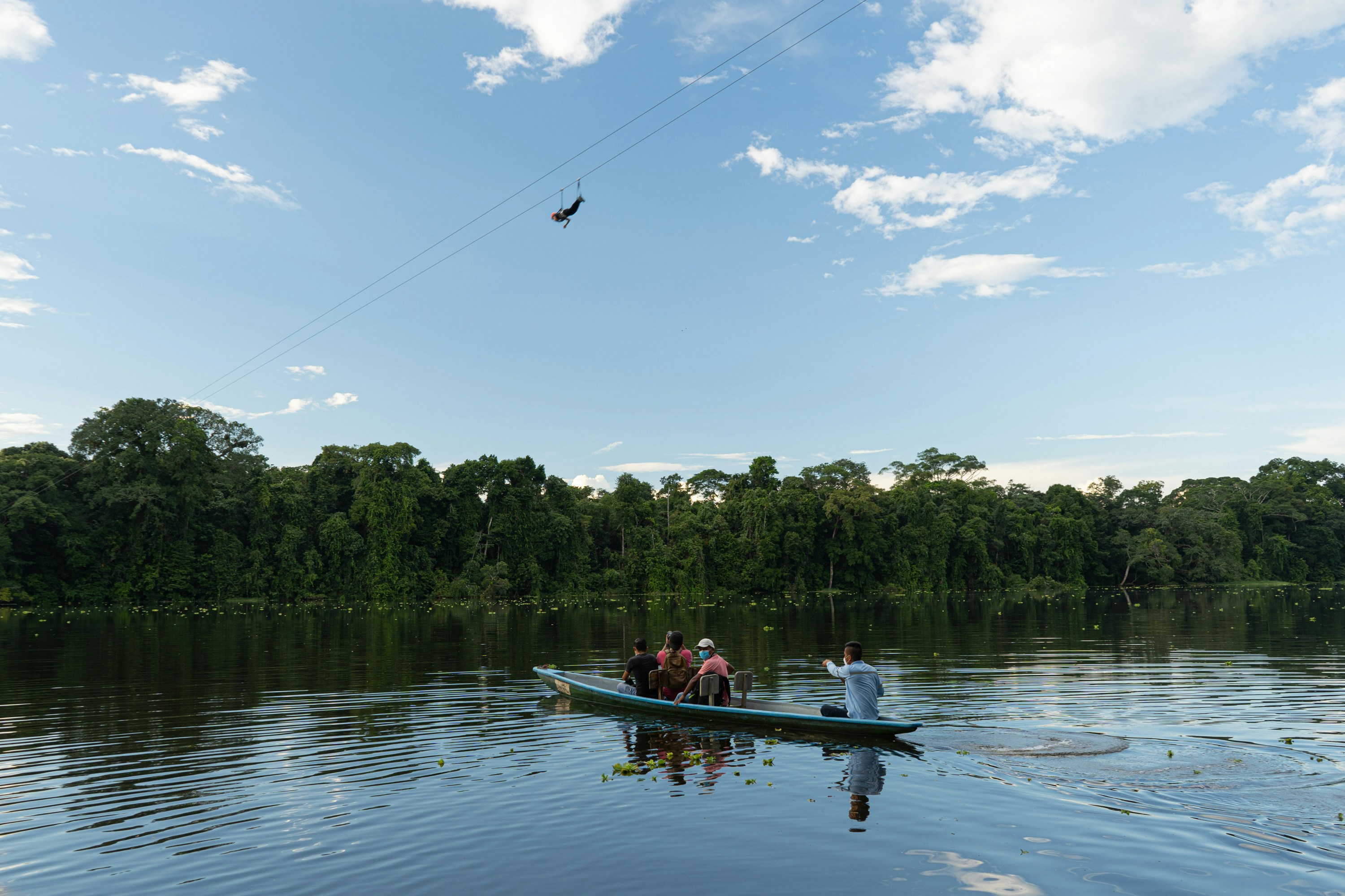 Group of people enjoying a boat ride on a tranquil river, surrounded by lush greenery and a zip-liner soaring above. 