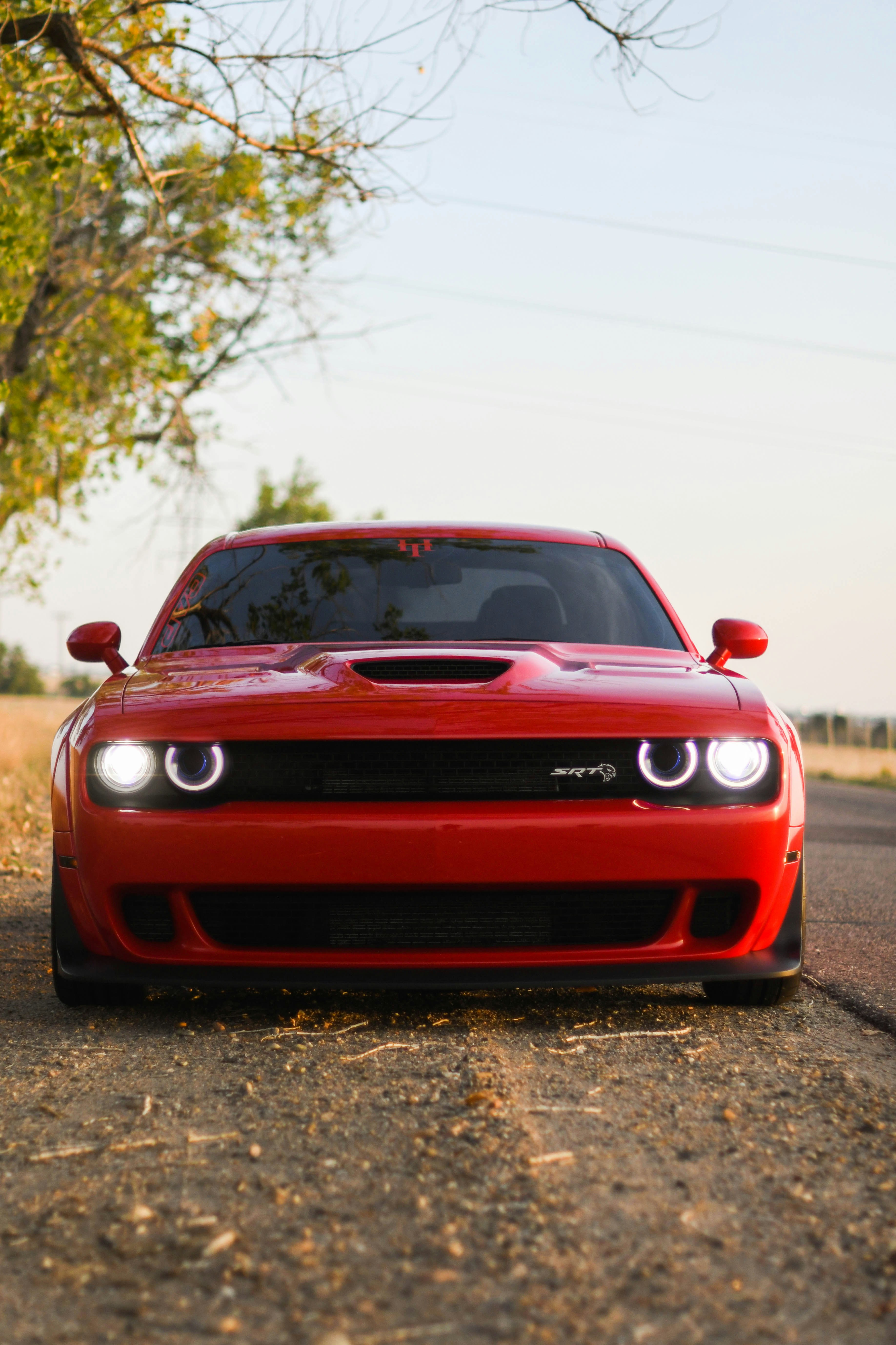 A striking red muscle car positioned on a rural road, showcasing its aggressive front design and distinctive headlights.