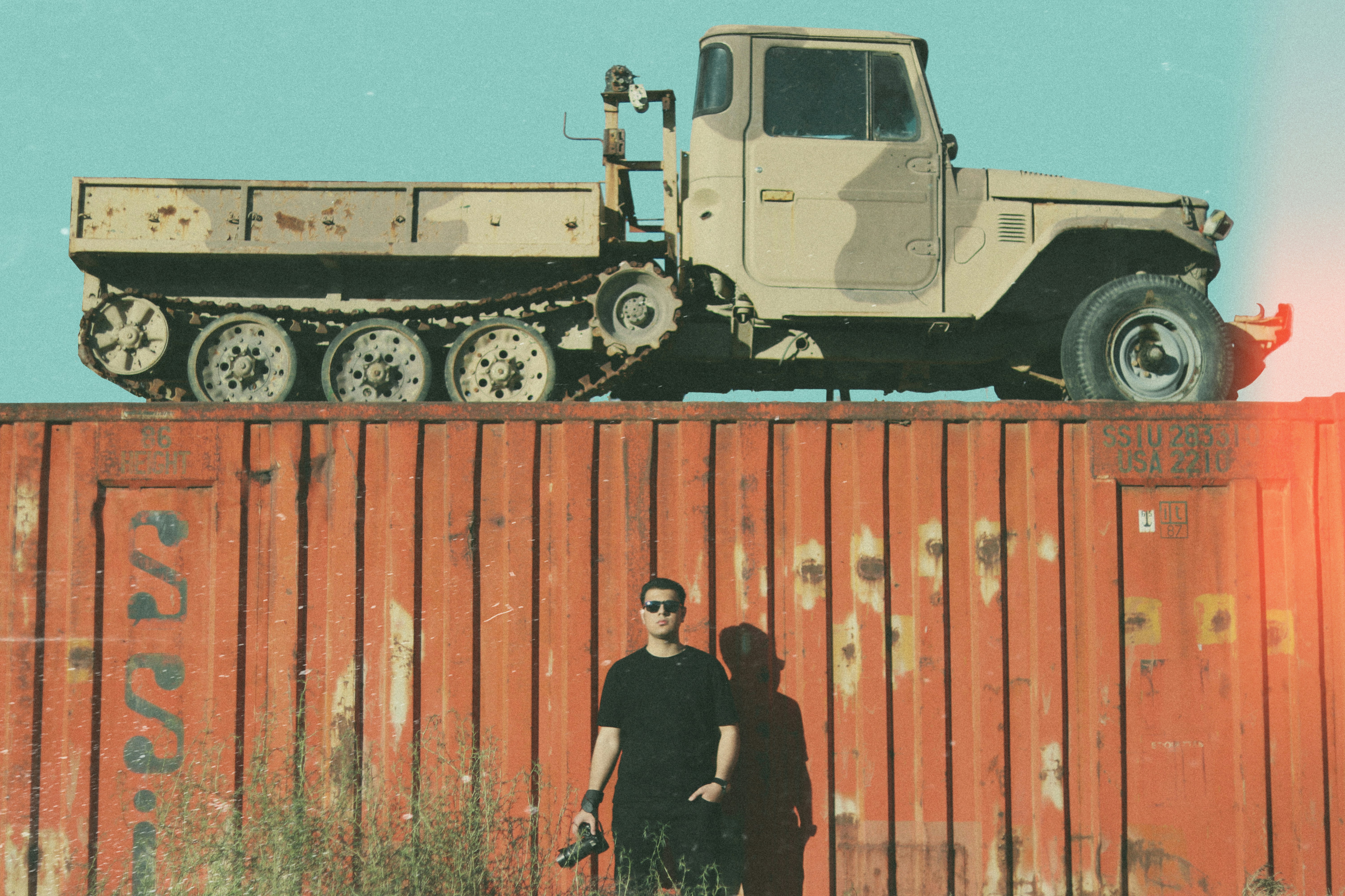 Man standing in front of a rusted red container with an old military vehicle on top.