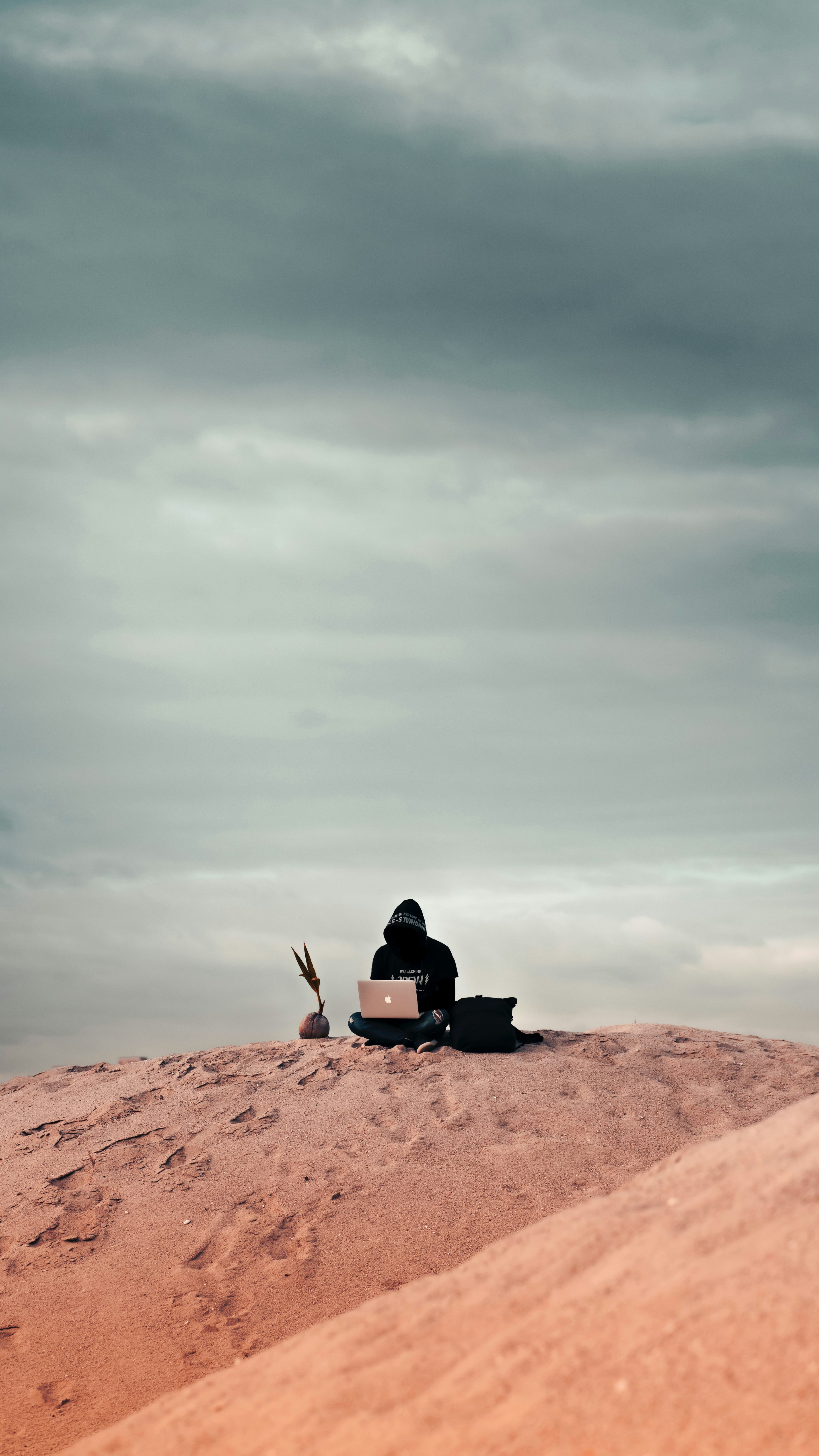 Individual working on a laptop atop a sandy dune, surrounded by minimalistic decor. A sense of solitude and focus permeates the scene.
