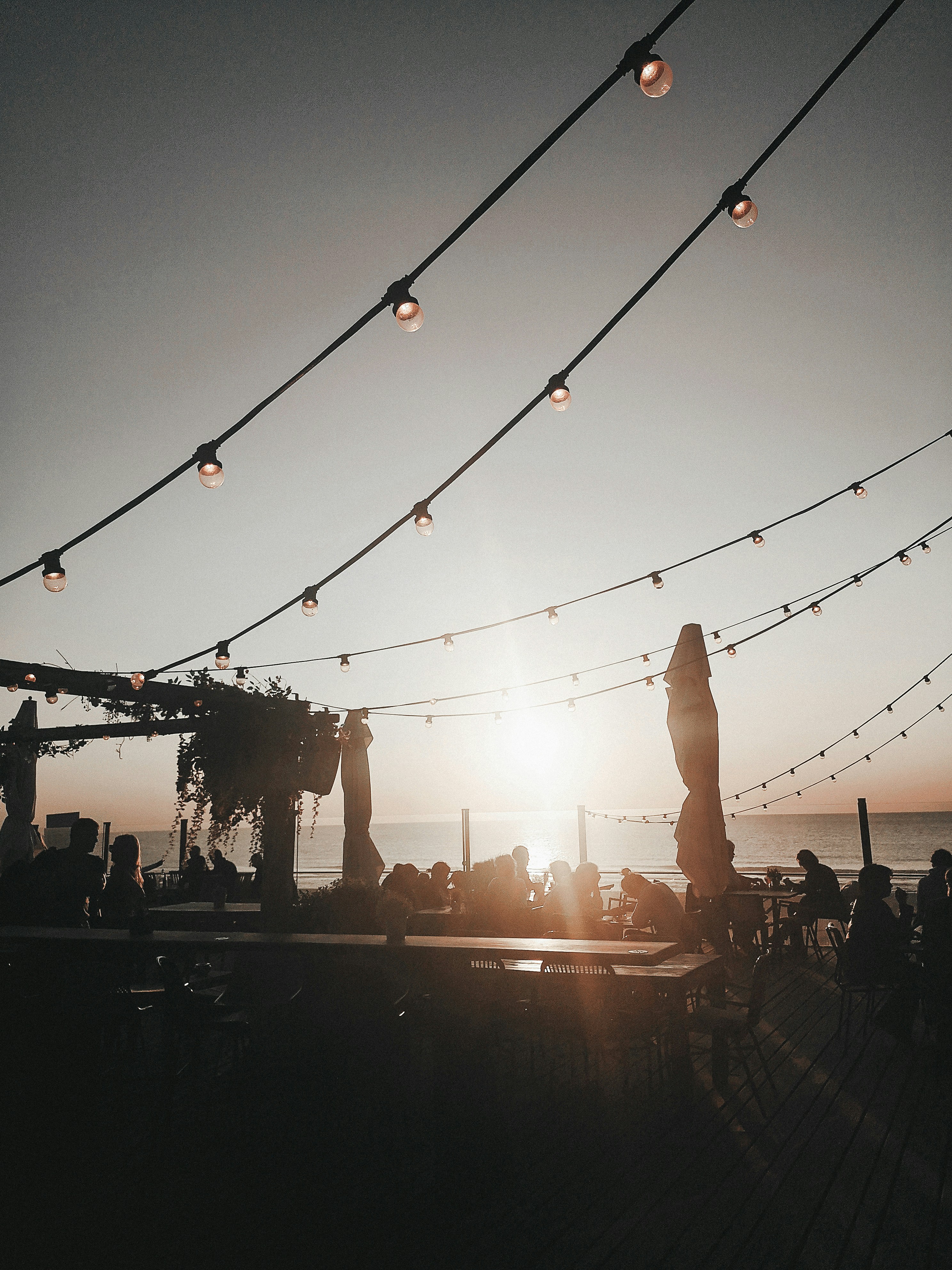 Warm sunset illuminating a lively beachside gathering with decorative string lights overhead. Guests enjoy the ambiance as the day transitions to night.