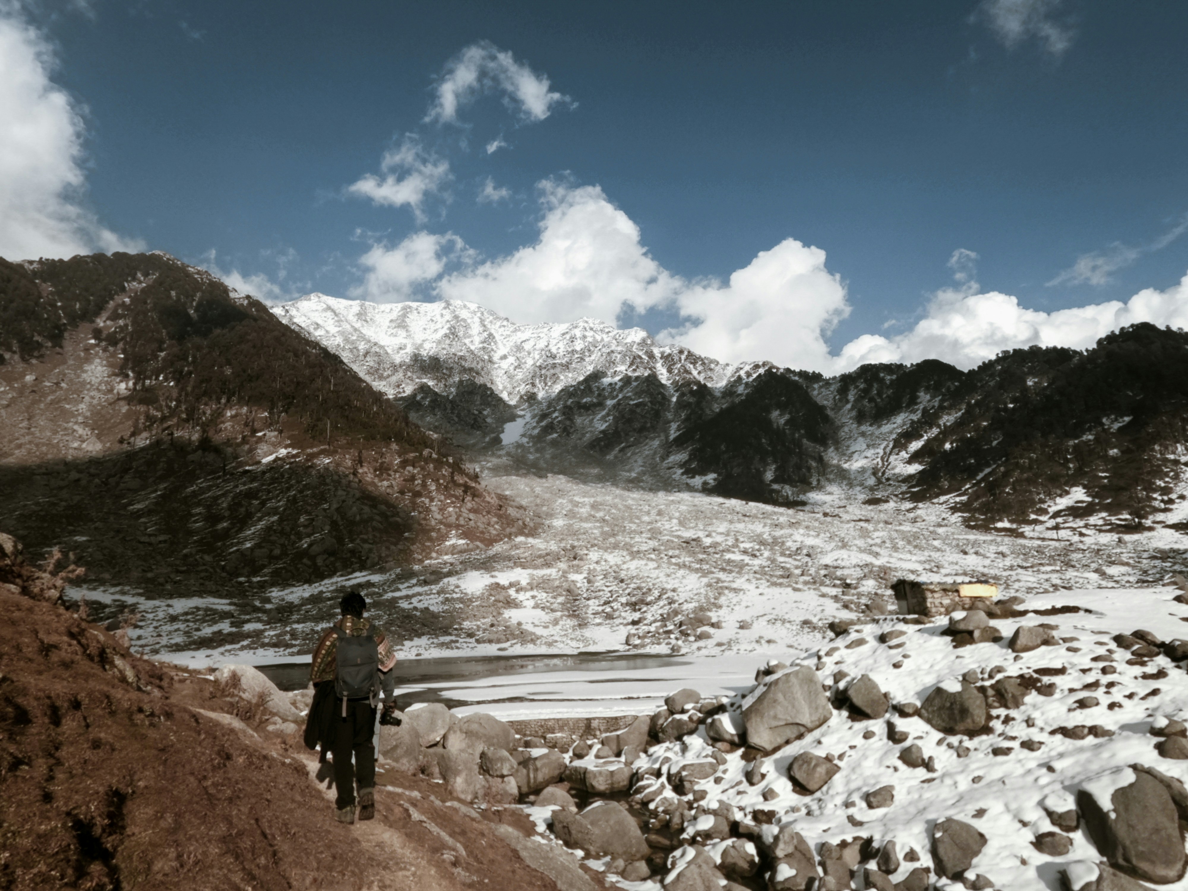 Hiker traversing a rocky path amidst snow-covered mountains under a dramatic sky.