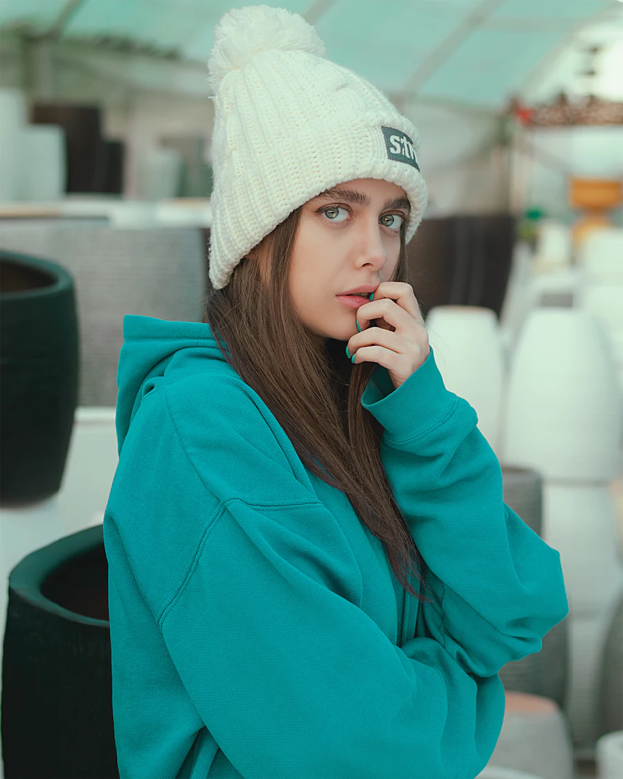 Young woman in a teal hoodie and white knit beanie looking toward the camera with her hand near her lips. Soft, blurred background with large pots or planters.