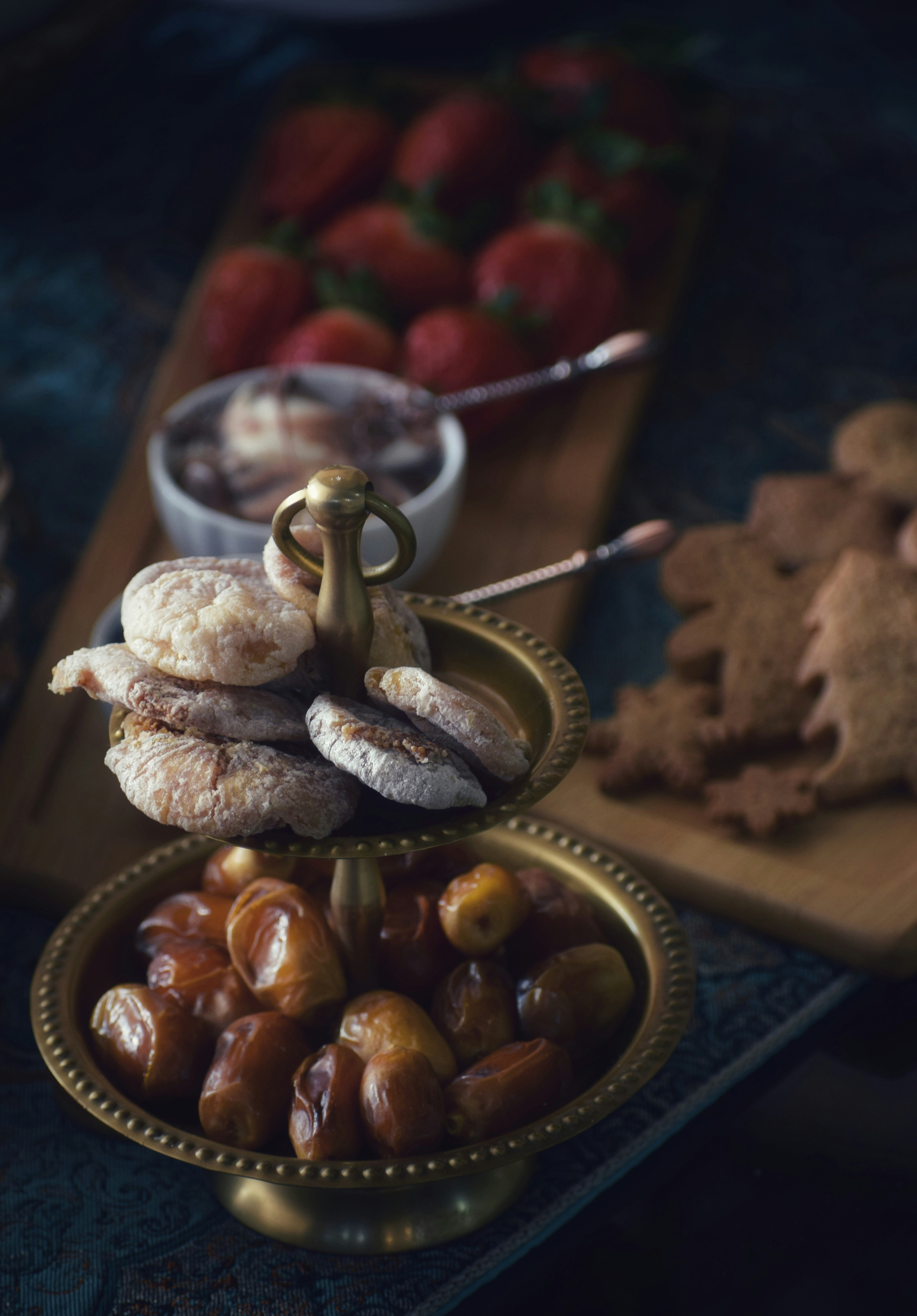 An elegant tiered stand displaying an assortment of cookies and dates, accompanied by fresh strawberries and a small bowl of chocolate sauce.