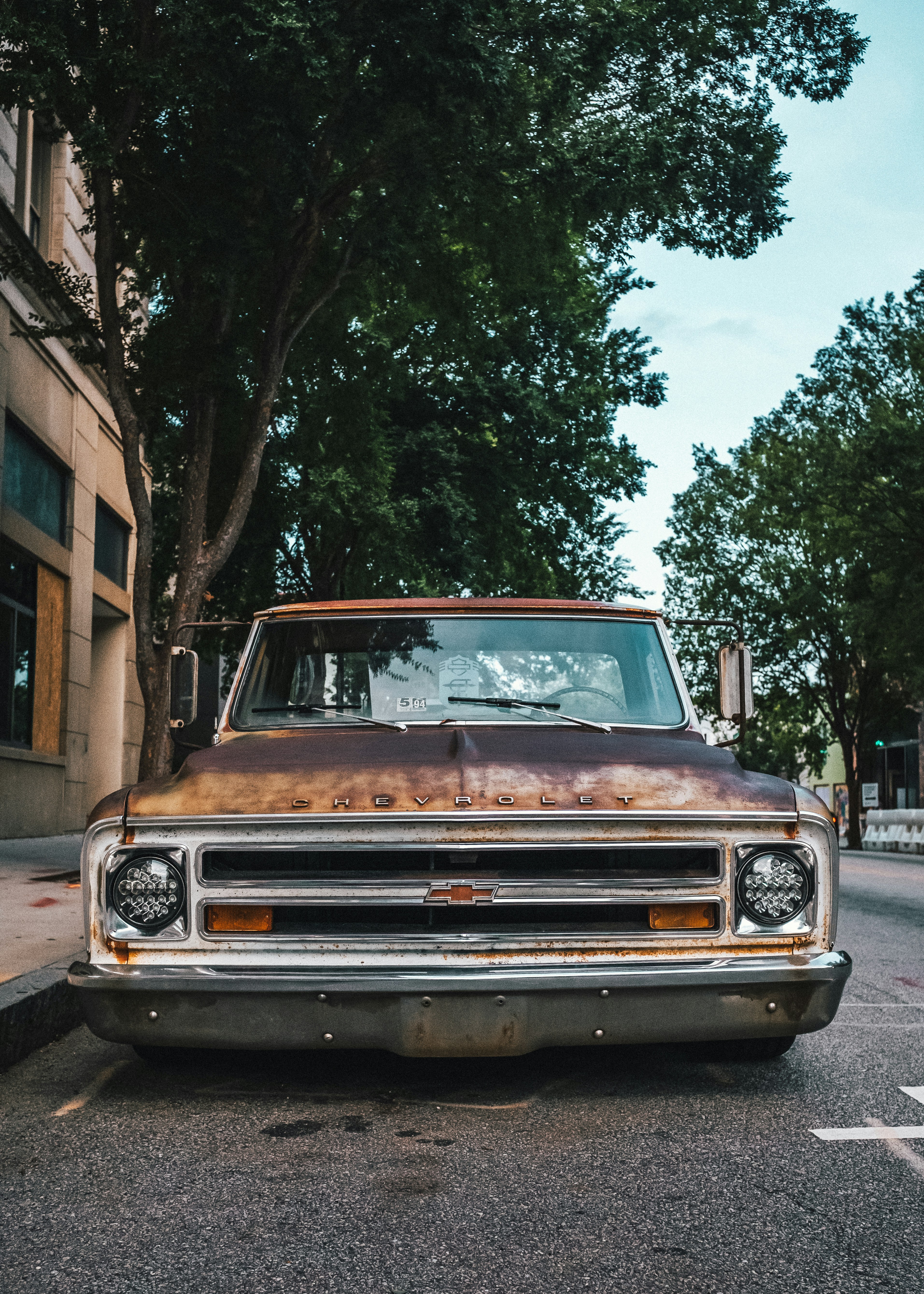 Weathered Chevrolet truck parked on city street, surrounded by lush trees and urban architecture.