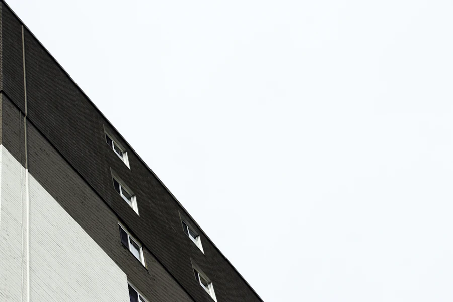 A dark apartment building facade cuts diagonally across the frame, with a row of small windows and a large expanse of pale sky. The strong geometric lines create a minimalist urban composition.