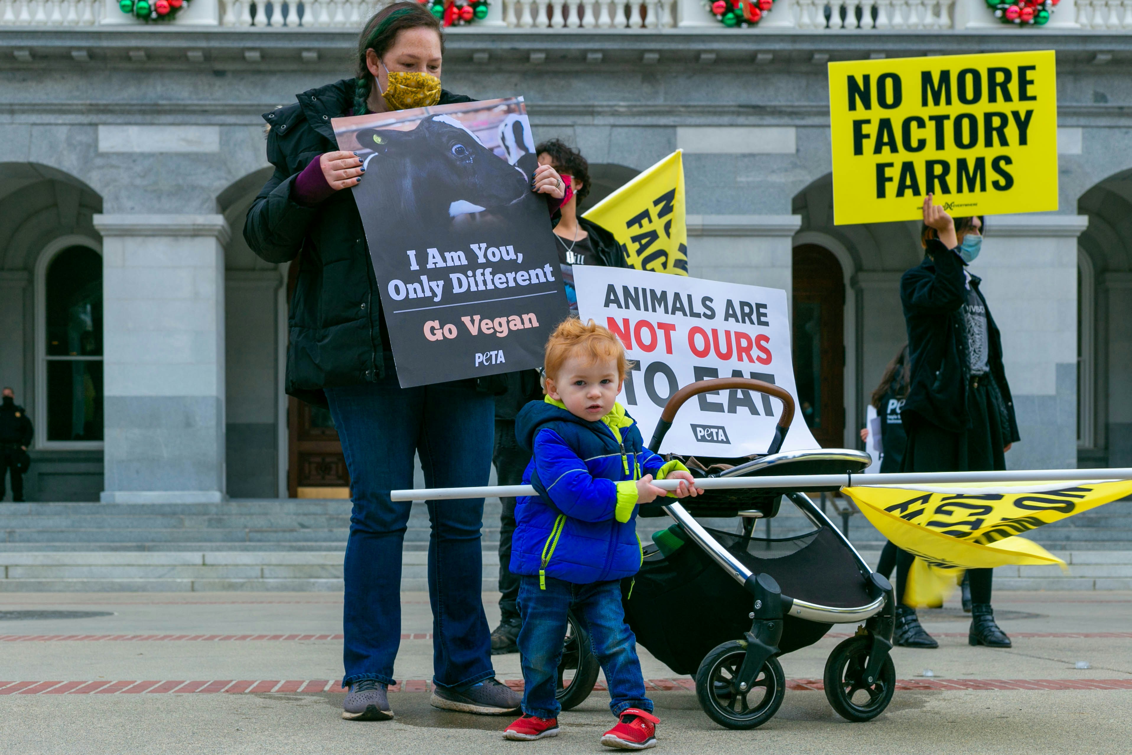 Demonstrators holding signs advocating for veganism and animal rights, with a child in a stroller at the forefront. The scene captures a moment of activism in an urban setting.