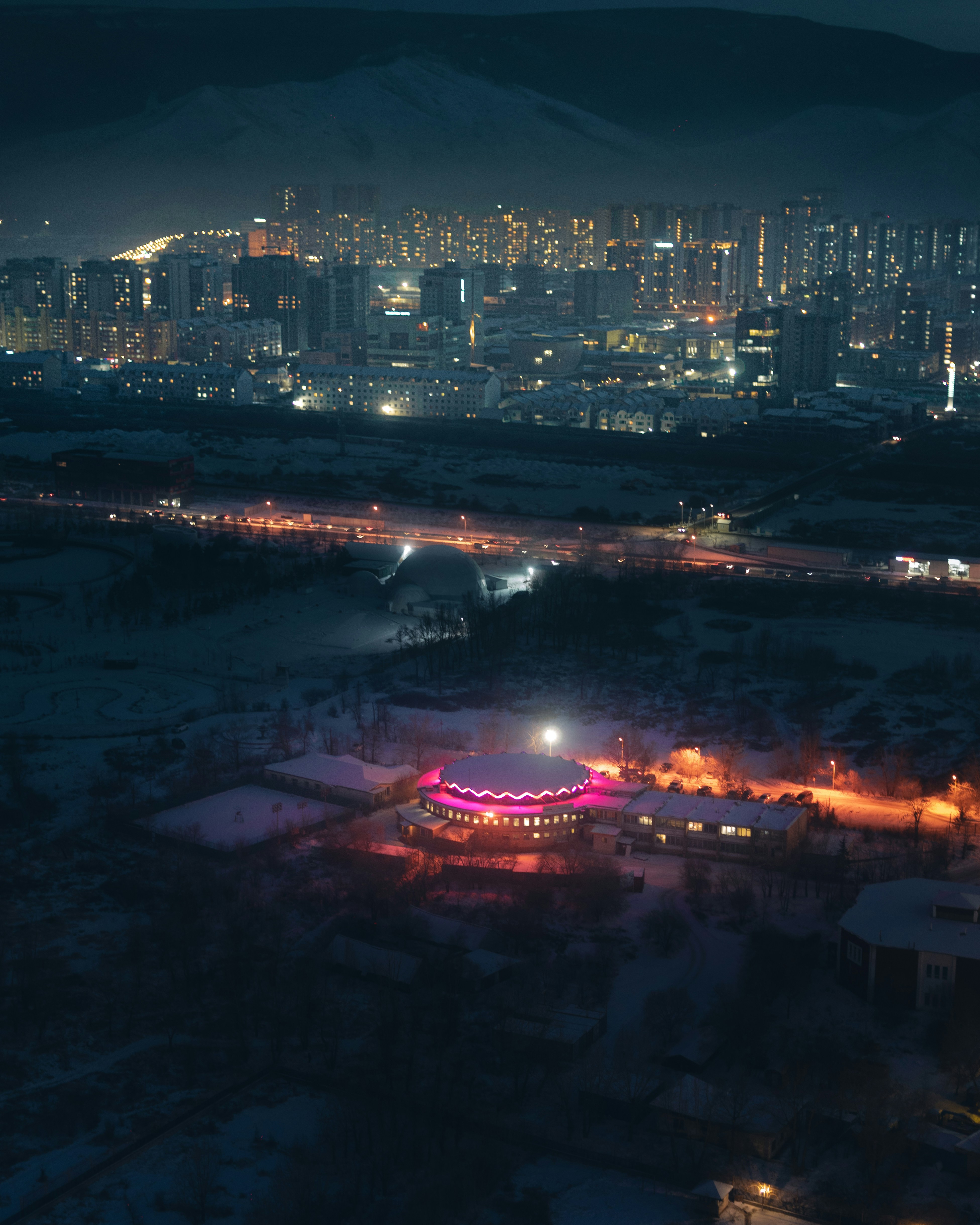 Vibrant circular structure illuminated in pink surrounded by a snowy landscape, with a sprawling city skyline in the background at night.
