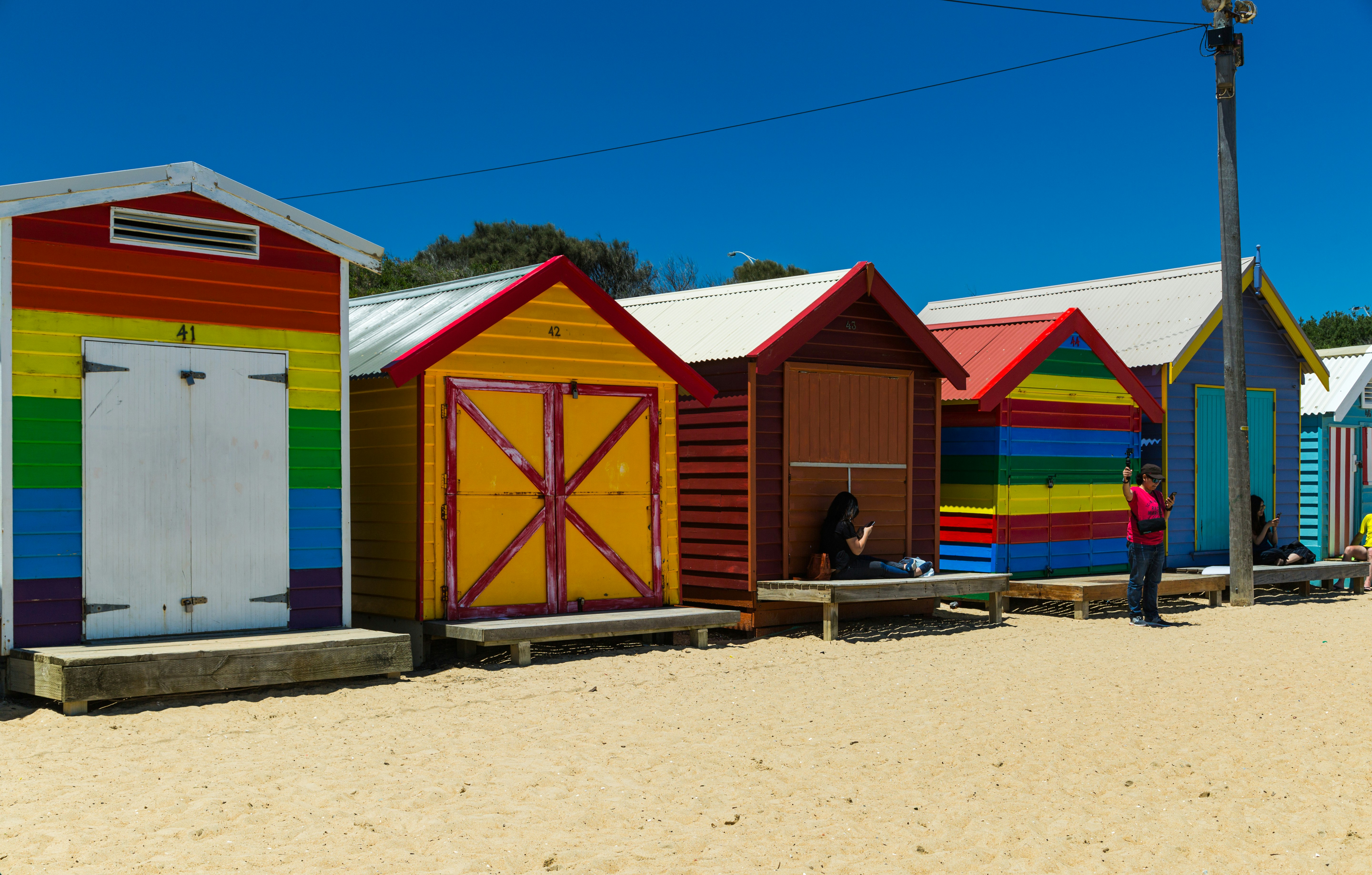 Colorful beach huts line a sandy shore, set against a bright blue sky.