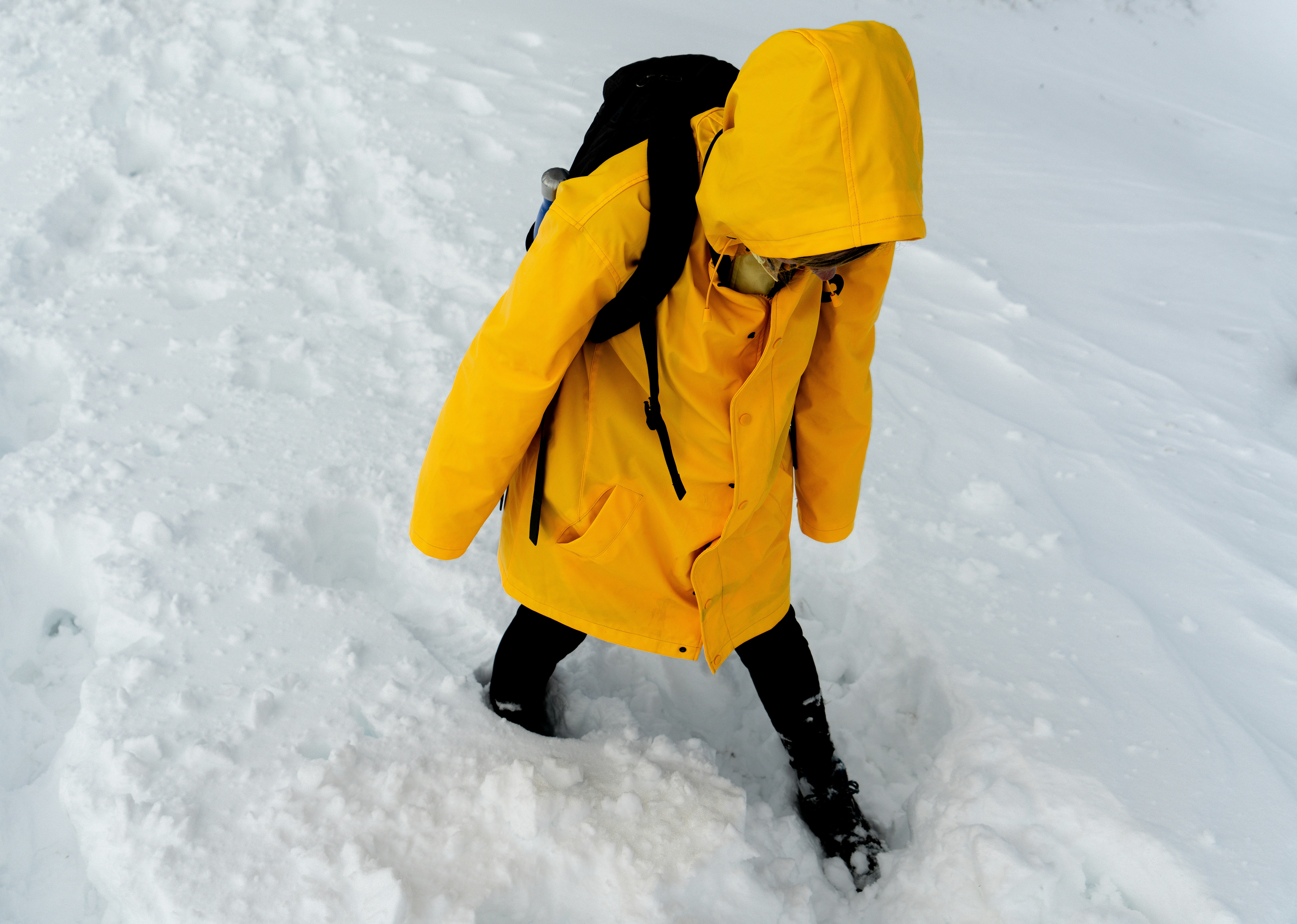 Person in a yellow coat trudging through deep snow with a black backpack.