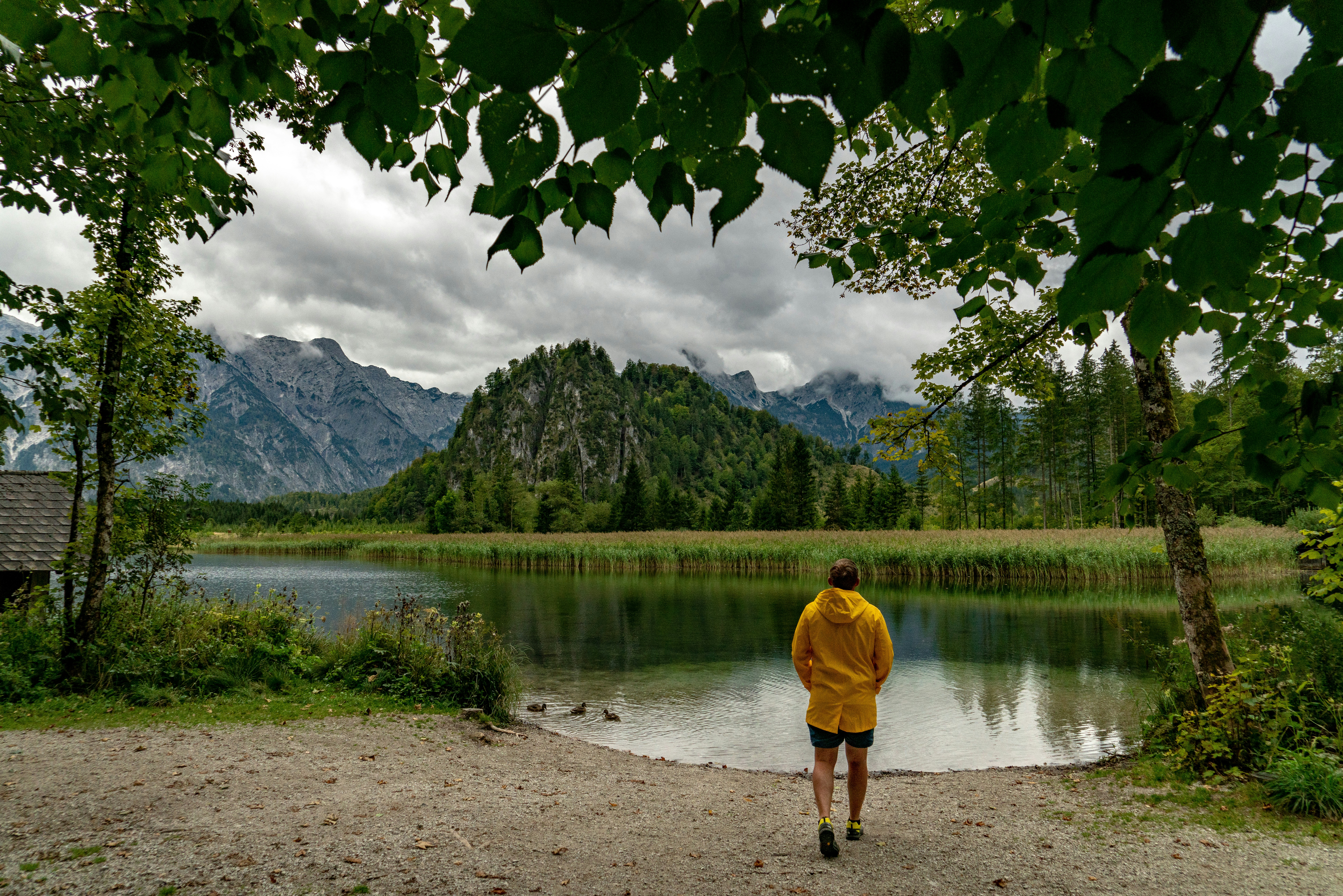 A lone figure in a yellow jacket stands by a tranquil lake, surrounded by lush greenery and majestic mountains under a cloudy sky.