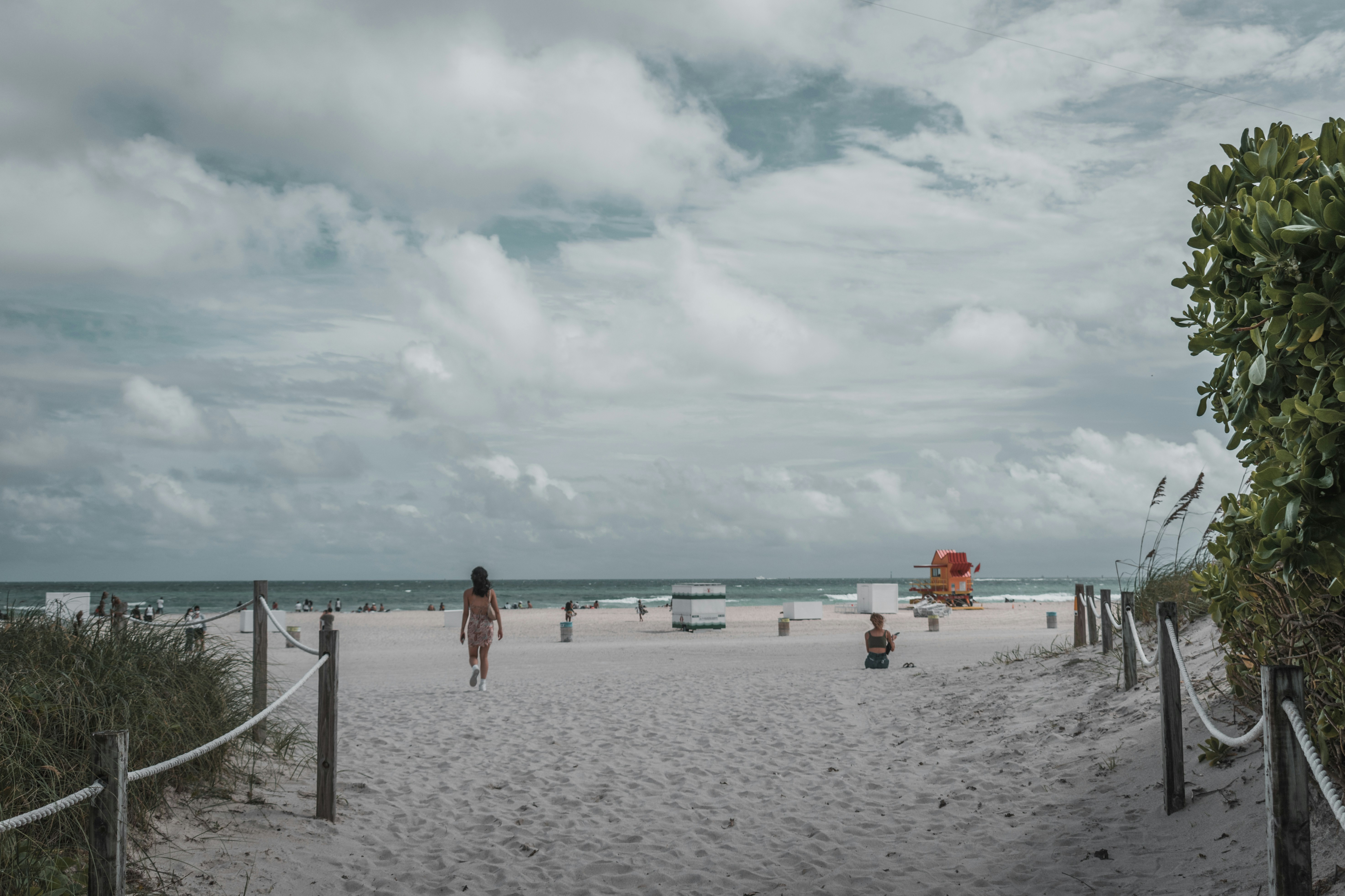 A tranquil beach scene with a pathway leading to the ocean, framed by lush greenery and scattered beachgoers. The sky is overcast, adding a serene ambiance.