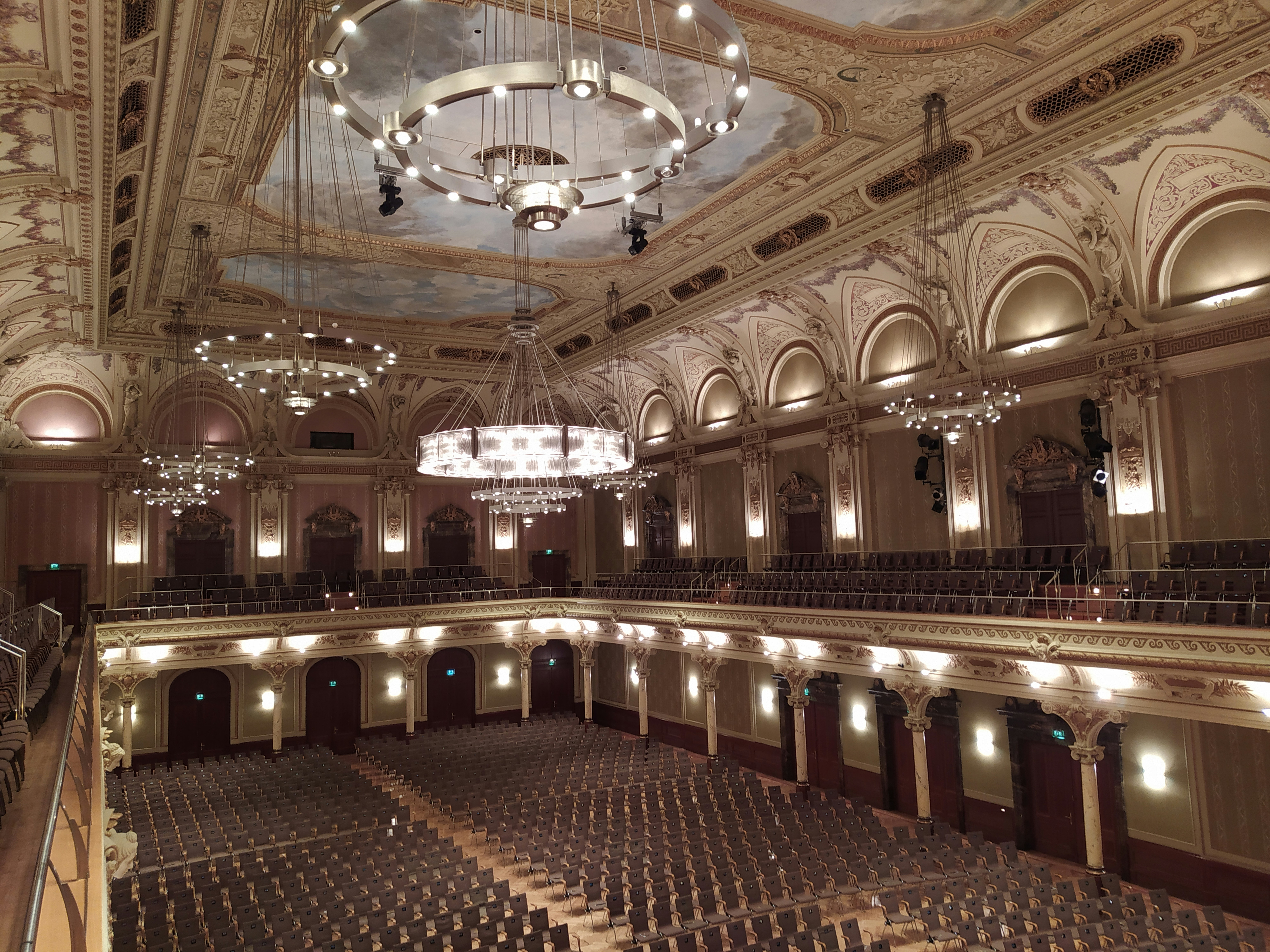 Elegant concert hall with ornate chandeliers and rows of brown wooden chairs.