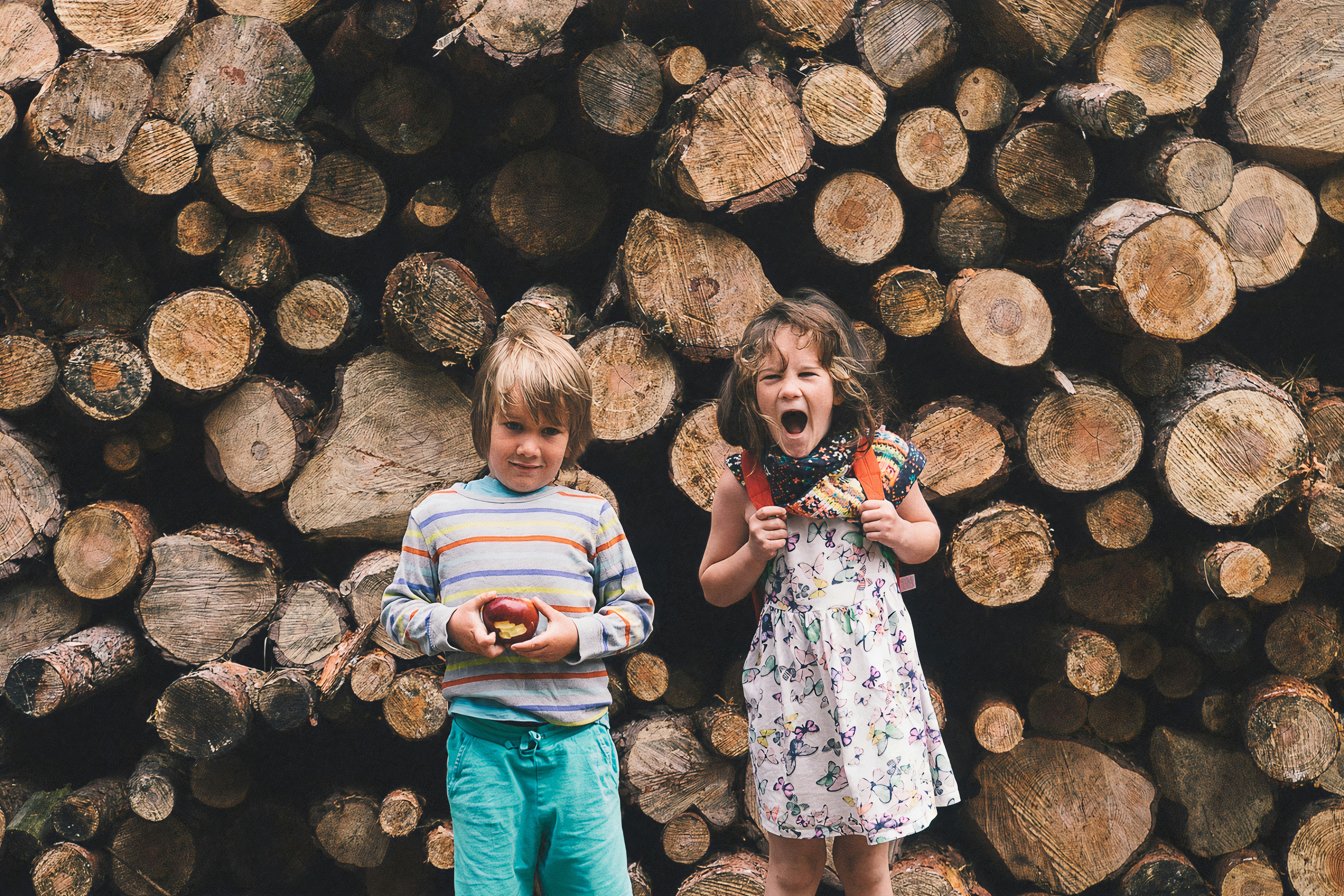 Two children stand in front of a stacked log backdrop, one holding an apple while the other joyfully poses with a toy. Their expressions embody the spirit of childhood adventure.