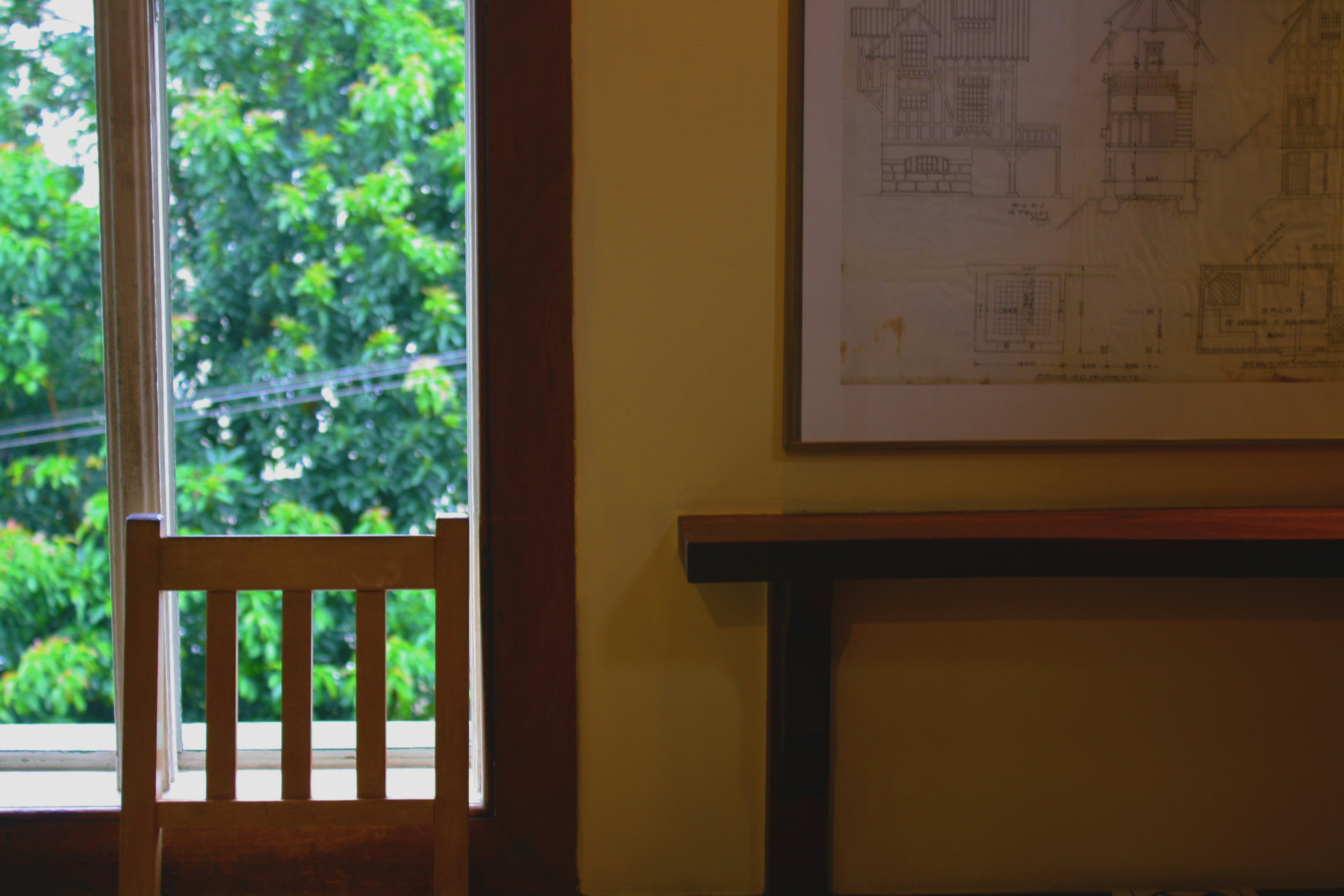 Wooden chair by a window overlooking lush greenery, framed by a soft yellow wall and a simple painting.