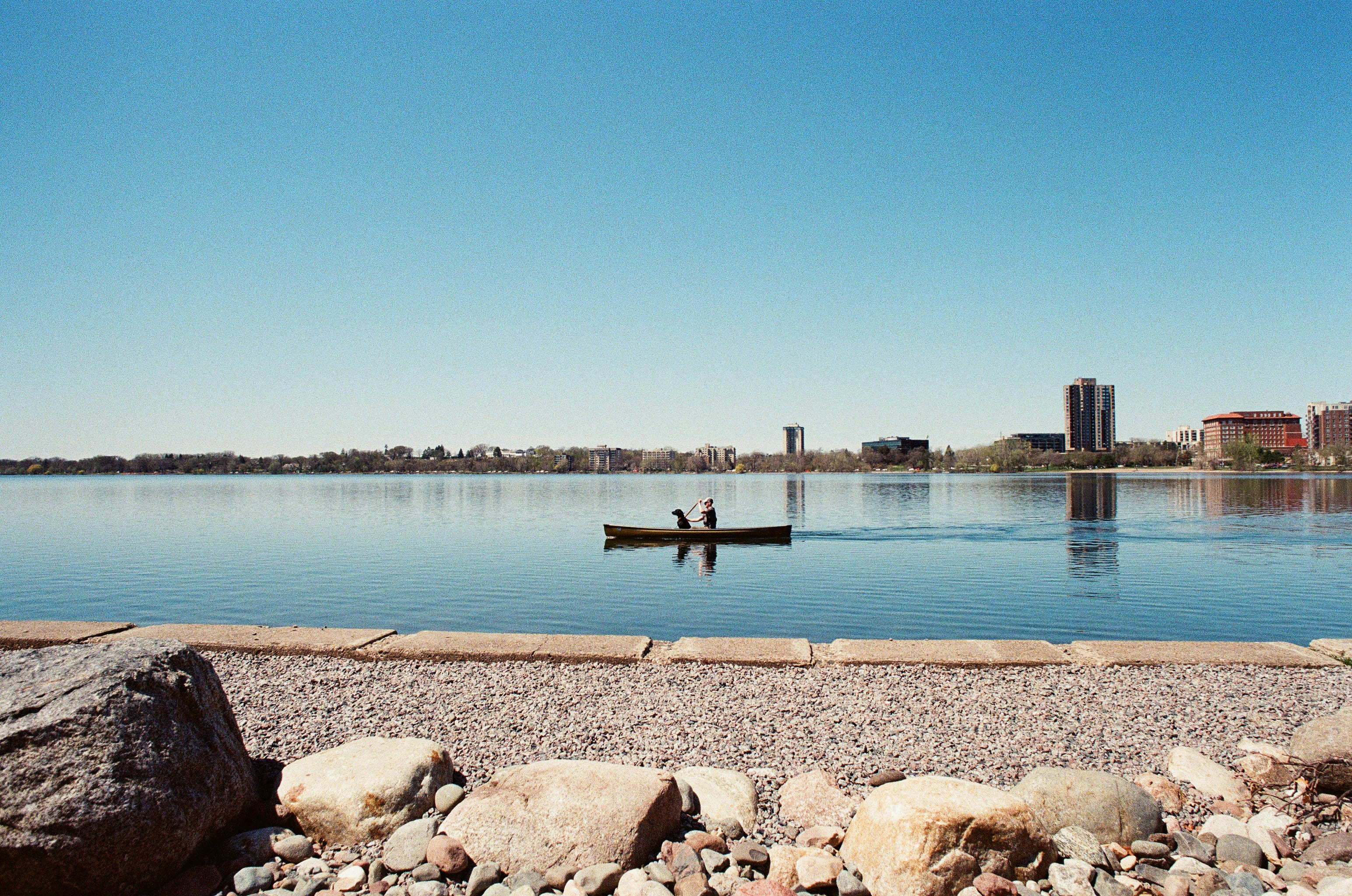 Canoe with two occupants gliding across a calm lake under a clear blue sky, with a distant cityscape on the horizon.