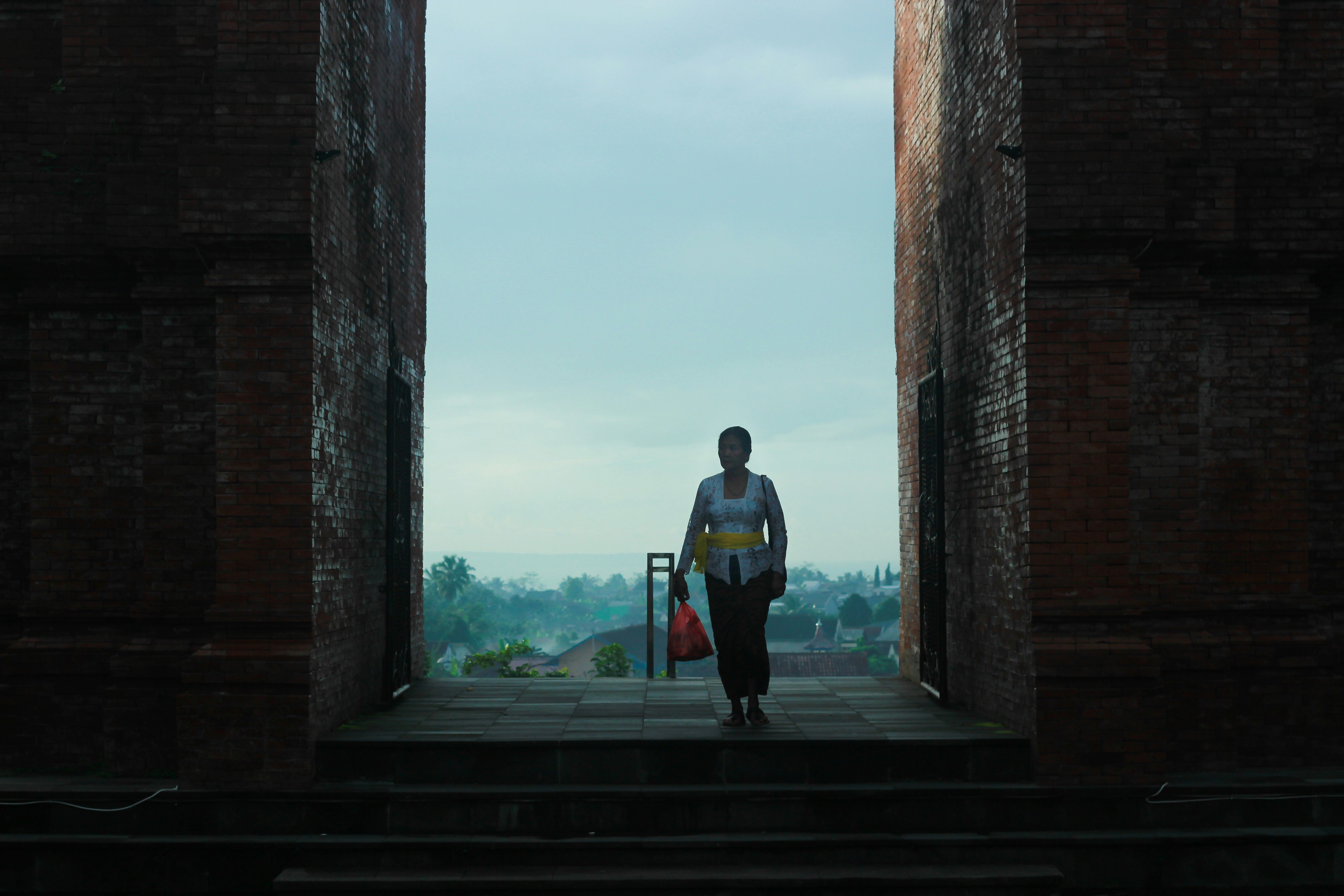 Person walking through a large stone gate with a distant landscape visible under a cloudy sky.