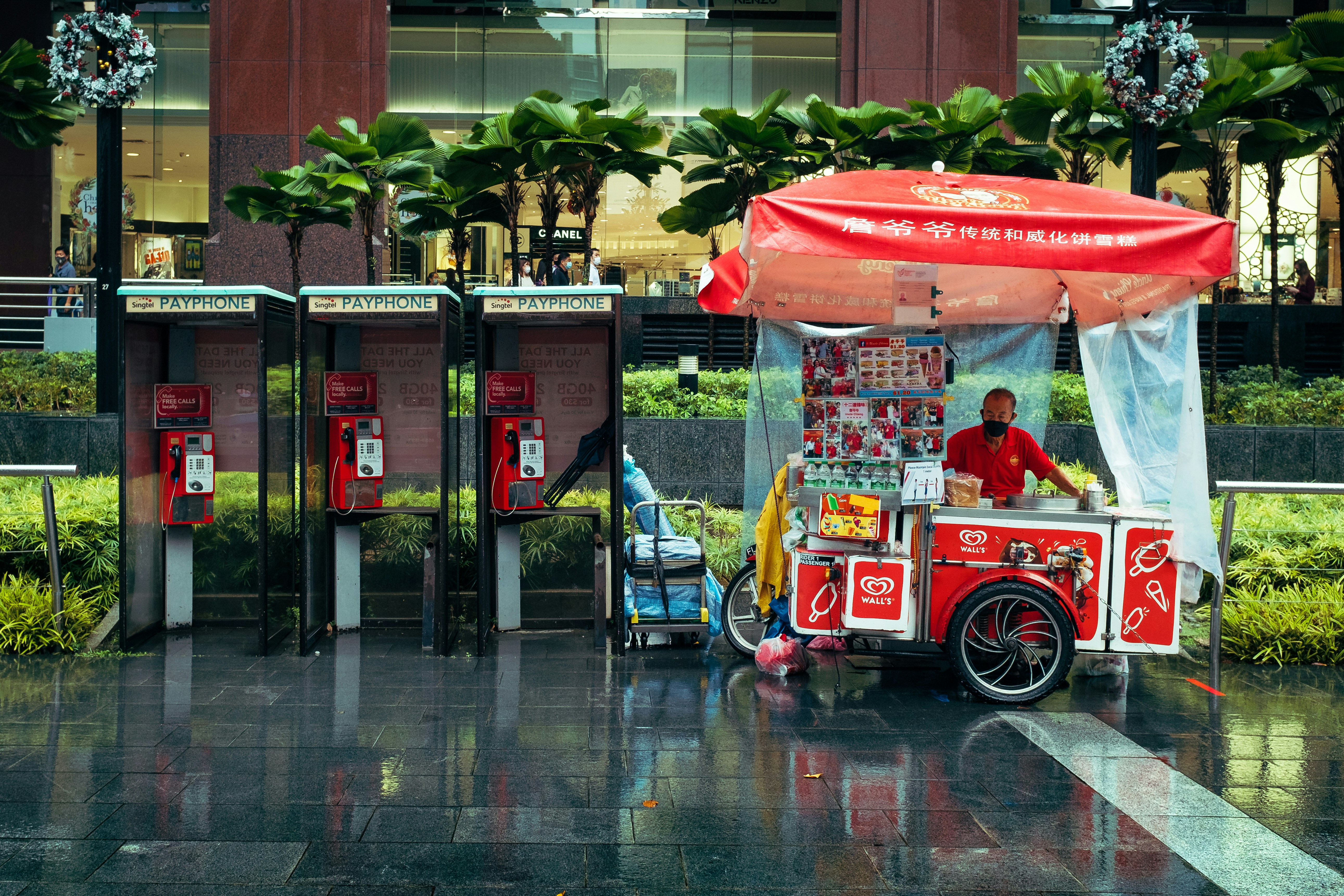 A vibrant food cart selling snacks under a red canopy, positioned in front of vintage payphones amidst lush greenery. Rain-soaked pavement reflects the lively scene.
