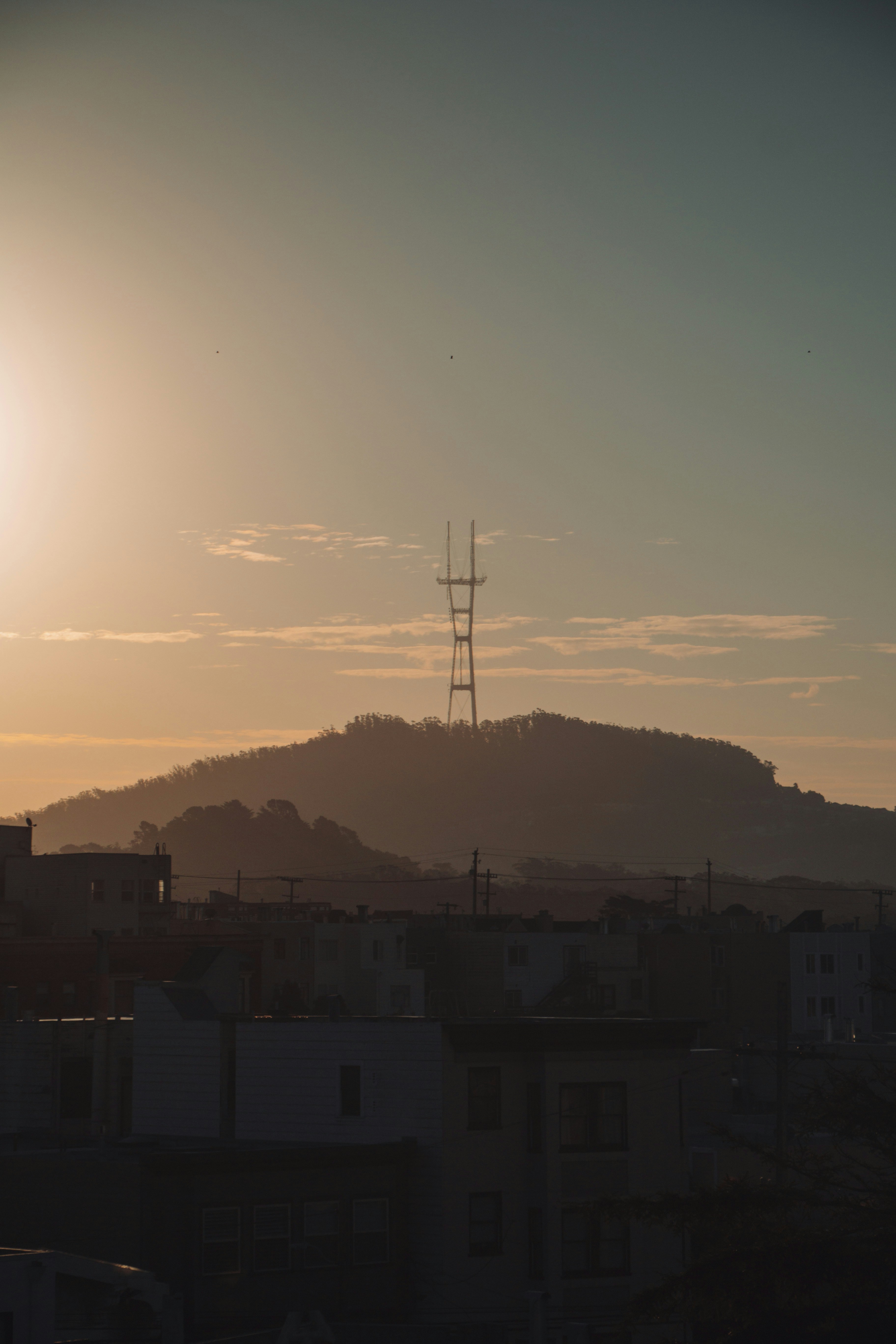 Telecommunication tower silhouetted against a soft sunset, with urban rooftops in the foreground. The scene captures the transition from day to night.