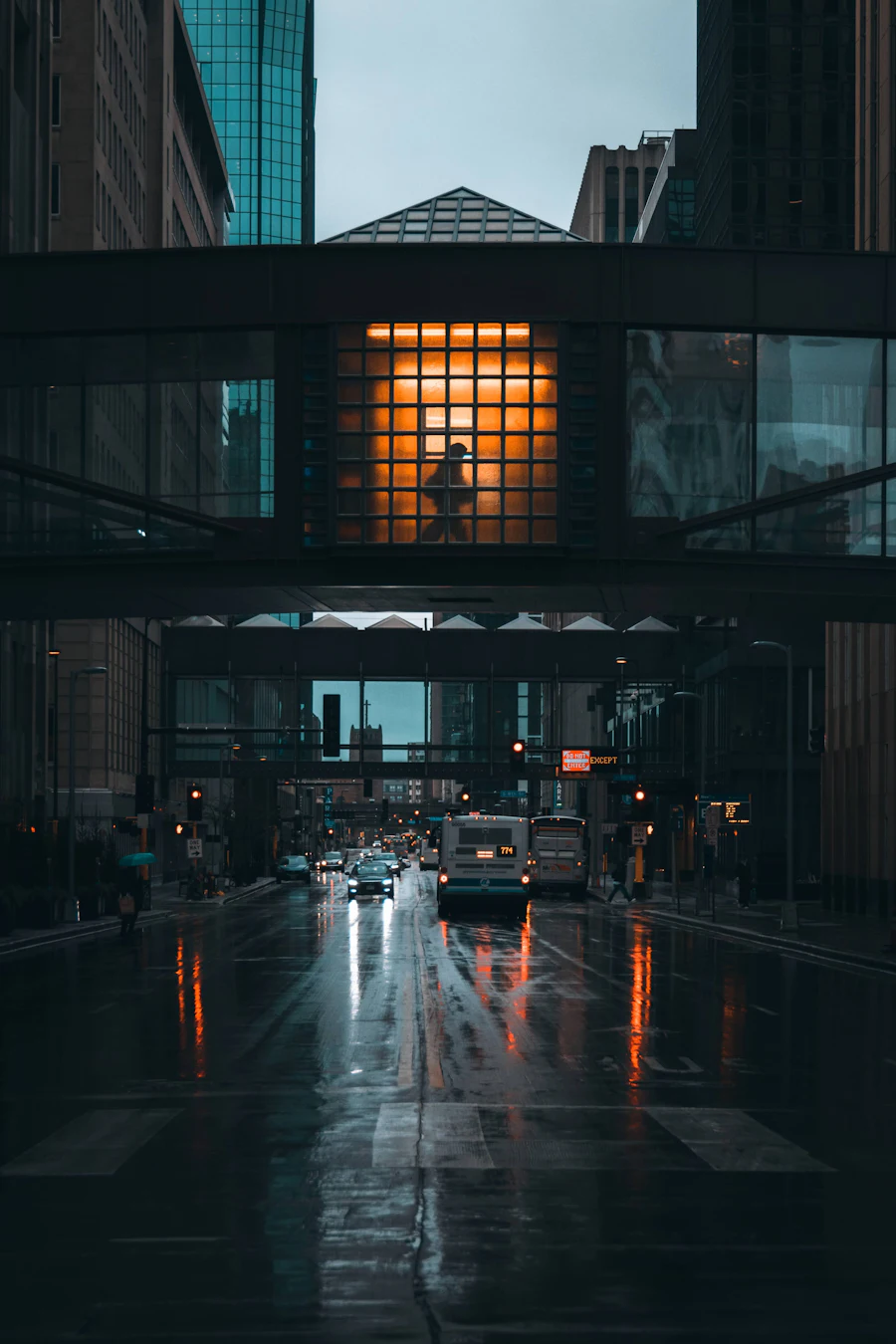 A wet downtown street framed by dark towers and an elevated skybridge, with a warm glowing window and a silhouetted figure above the road. Headlights and streetlights streak reflections across the slick pavement.