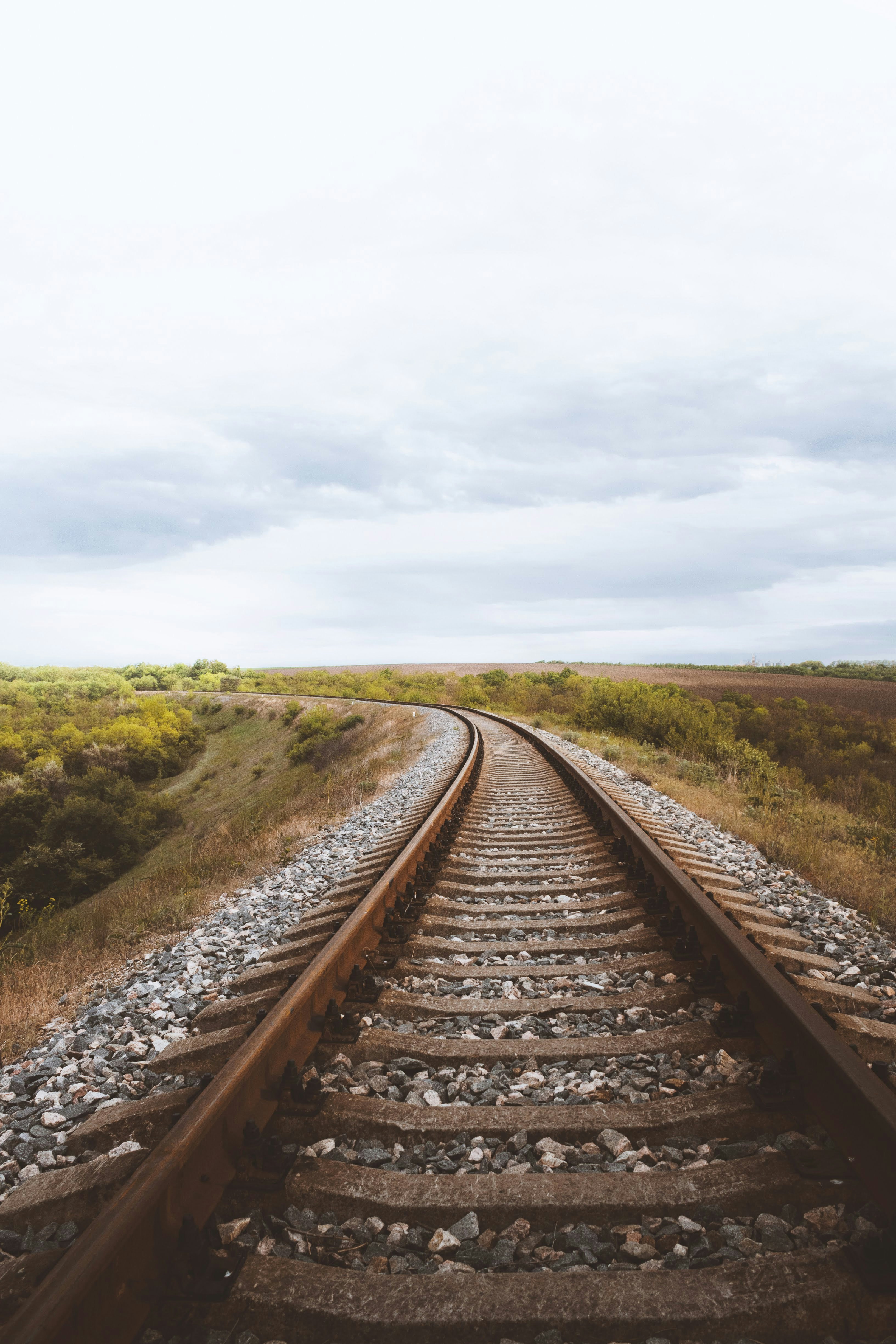 Curved railway tracks stretch into the distance, bordered by lush greenery and open fields under a cloudy sky.