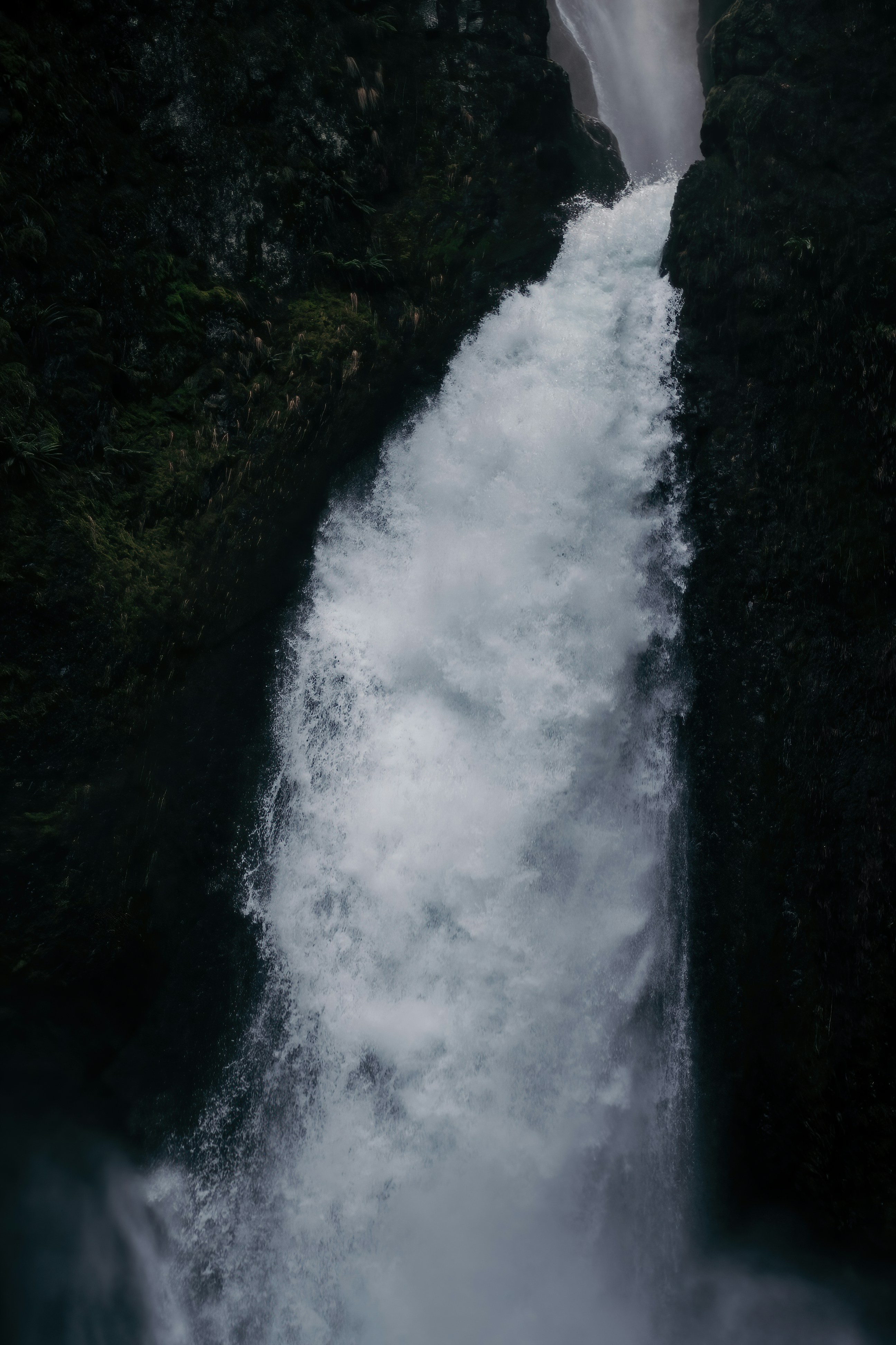 Rushing waterfall cascading through a narrow gorge, surrounded by lush greenery and rocky cliffs.