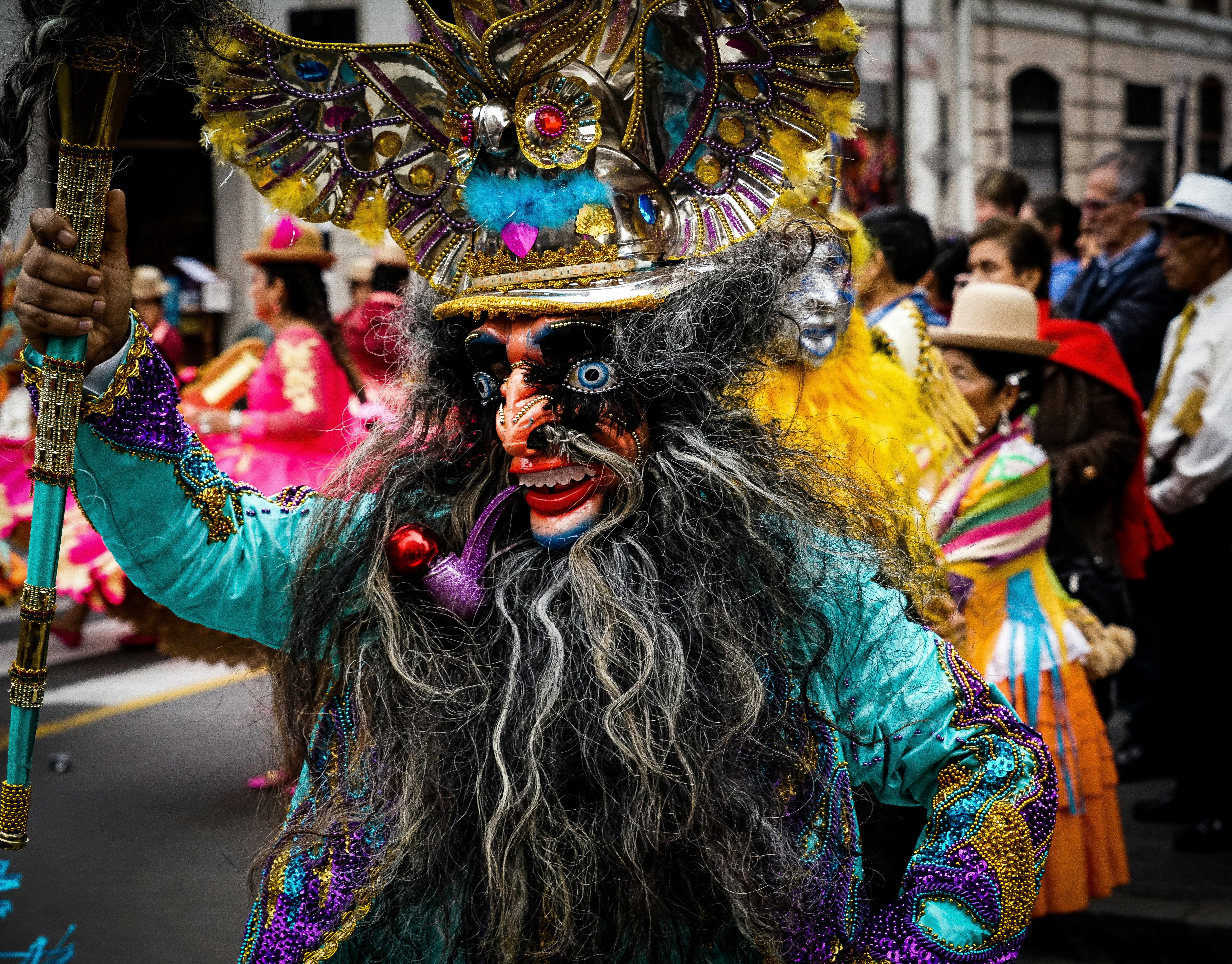 A colorful figure adorned in elaborate costume and makeup, embodying the spirit of a lively festival, surrounded by a backdrop of joyful participants in traditional attire.