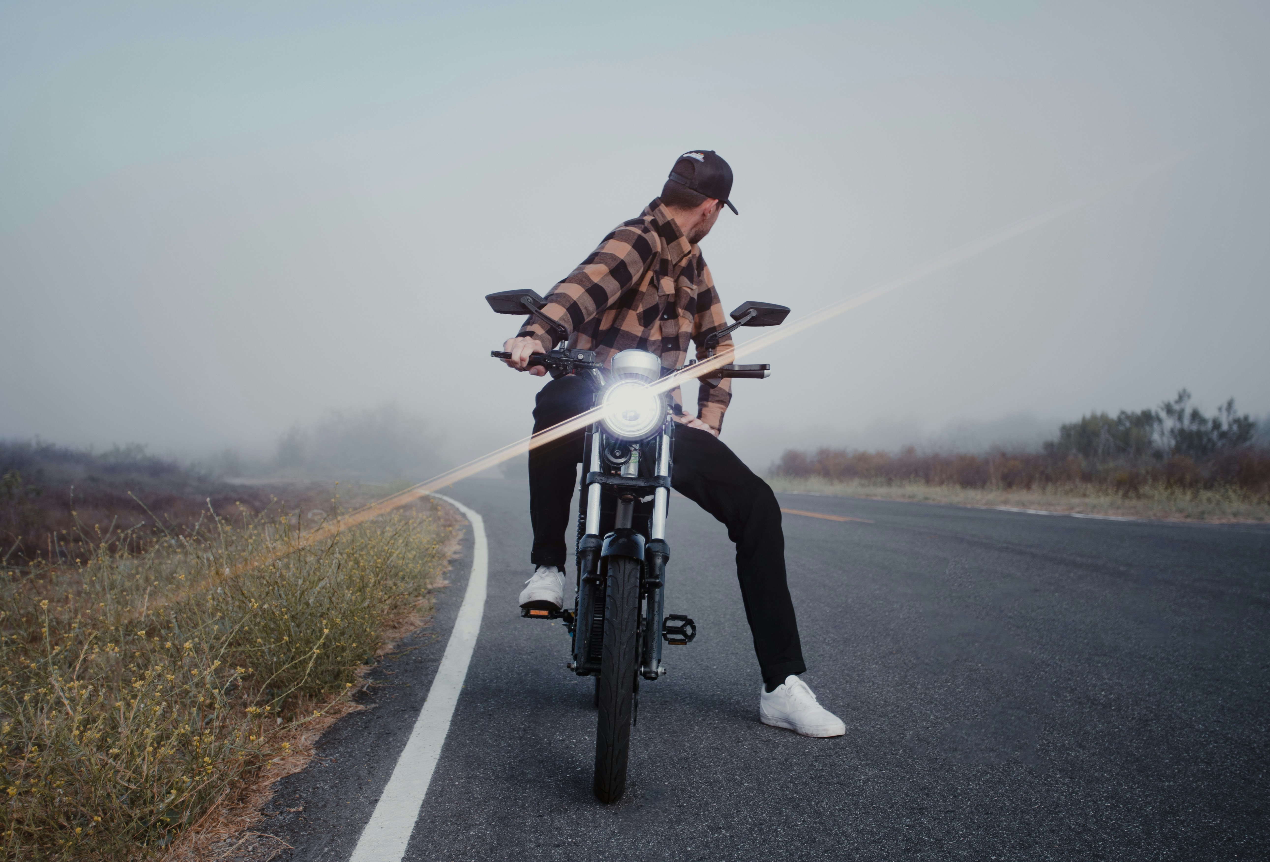 A person in a plaid shirt rides a motorcycle on a winding road enveloped in fog, creating an atmosphere of adventure and mystery.