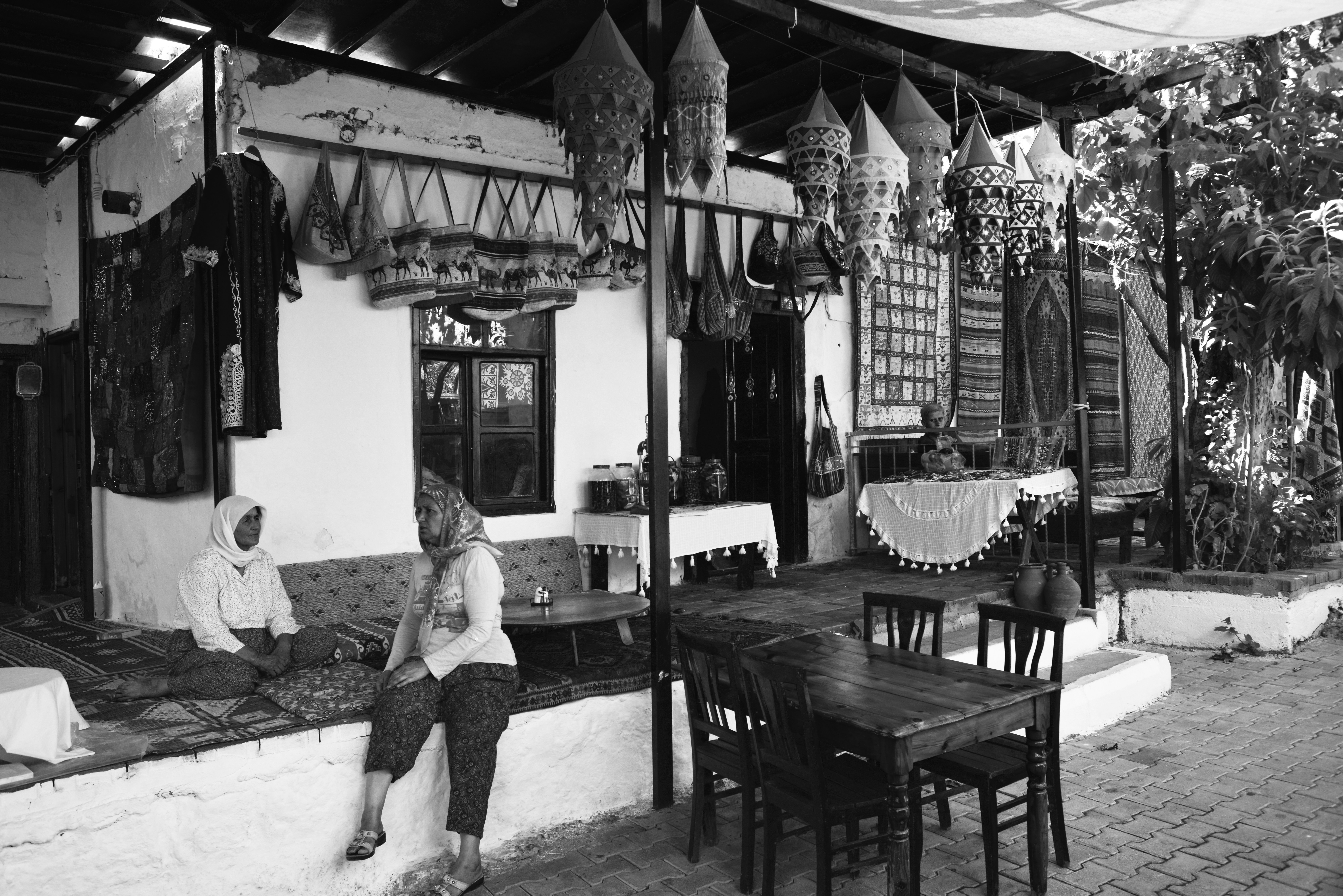 Two women engaged in conversation in a quaint outdoor setting adorned with handmade textiles and lanterns. The scene reflects cultural heritage and community spirit.