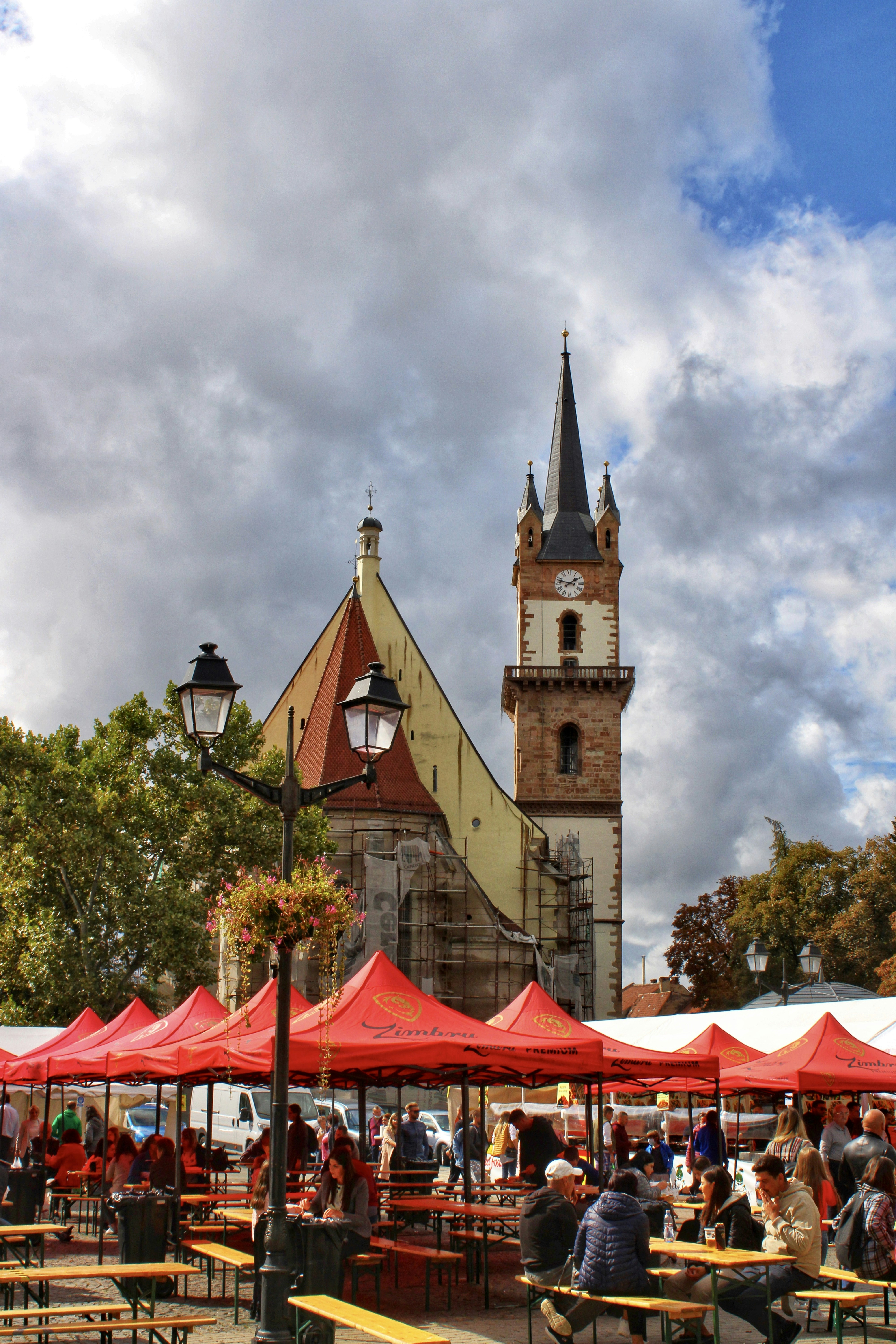 Bustling market scene with colorful tents and people enjoying the day, framed by a historic church tower and dramatic cloudscape.