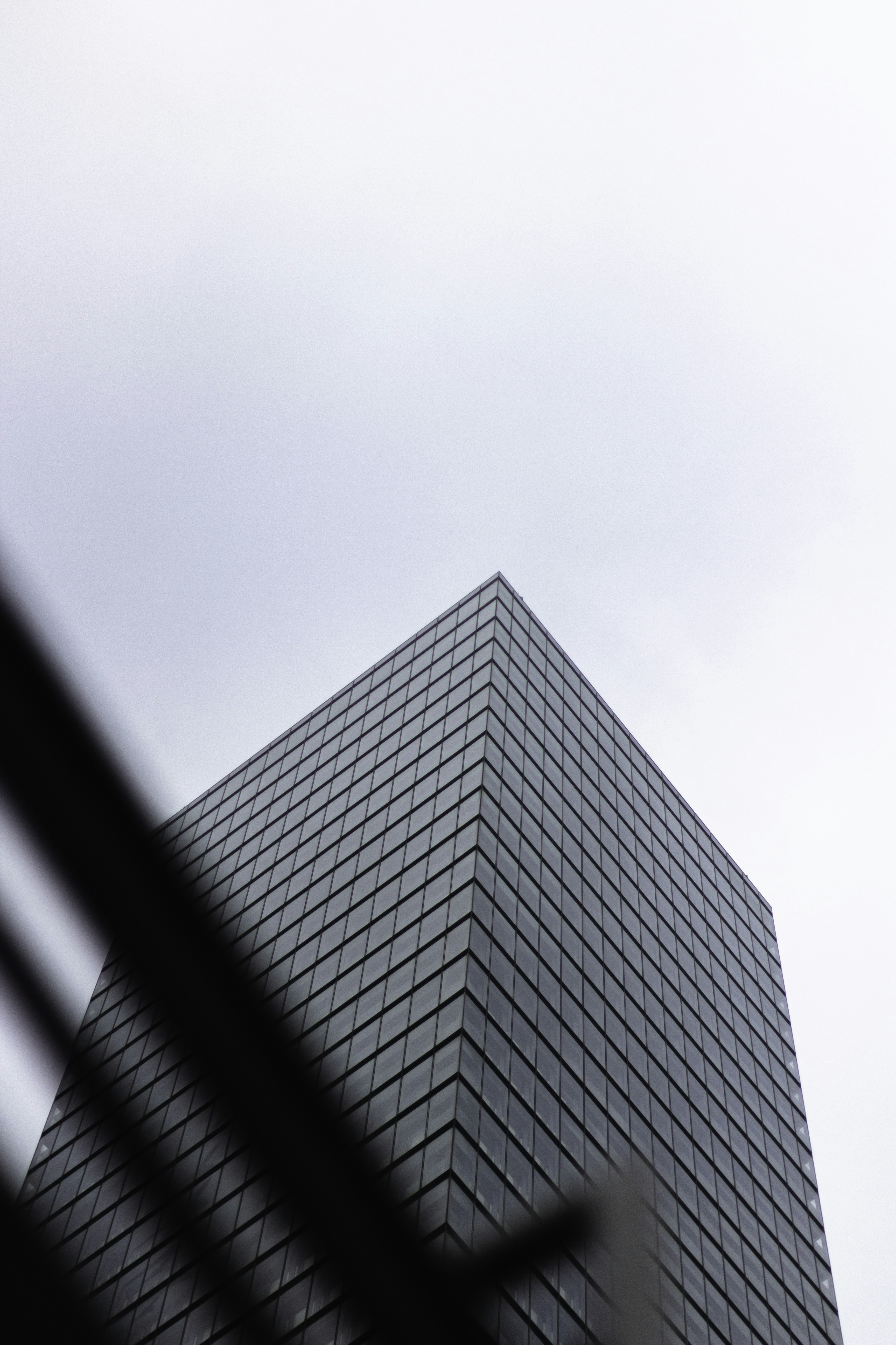 A modern skyscraper with a glass facade reaching towards an overcast sky, framed by blurred architectural elements in the foreground.