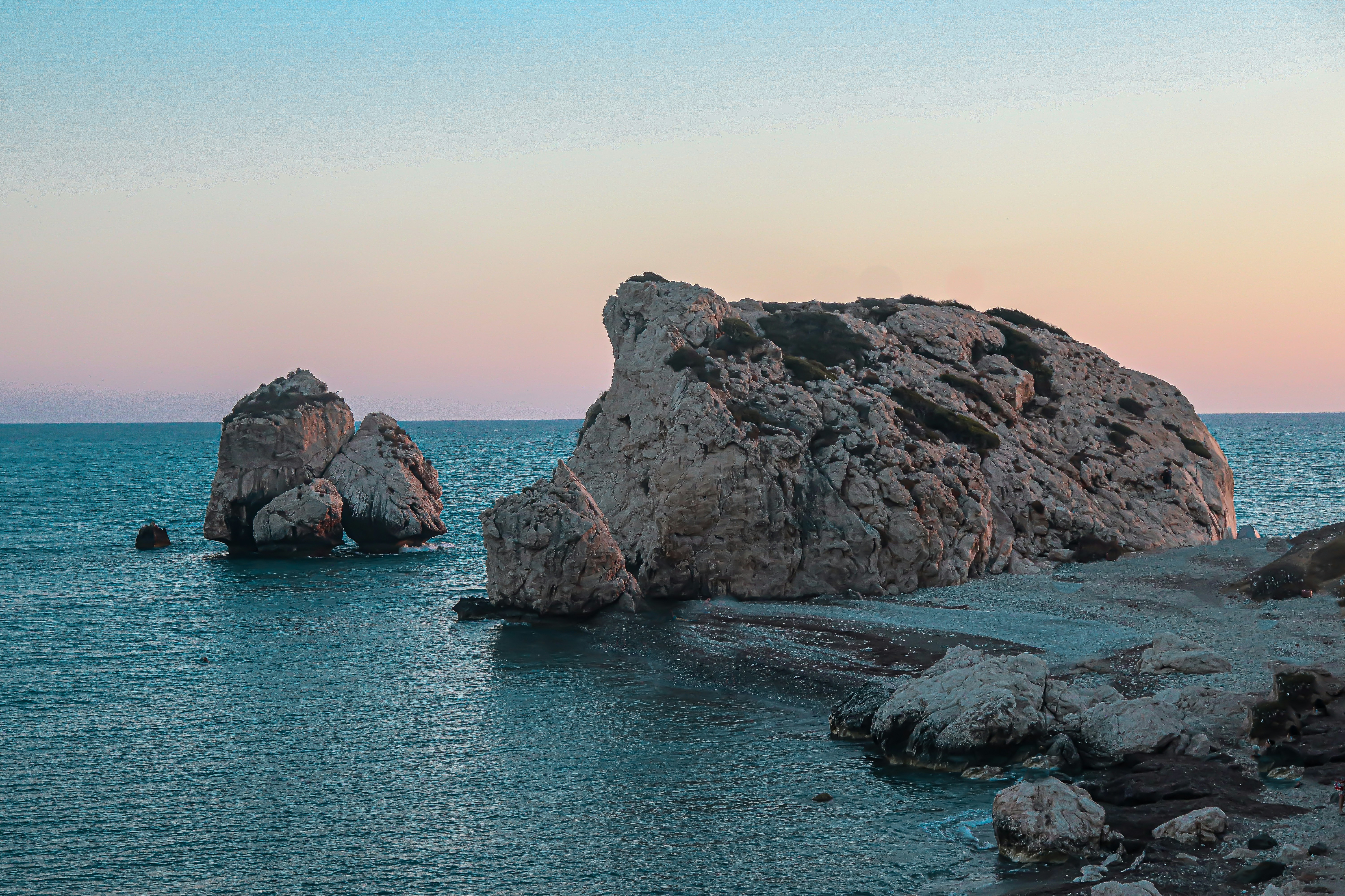 Large rock formations in a calm blue sea under a pastel sunset sky.