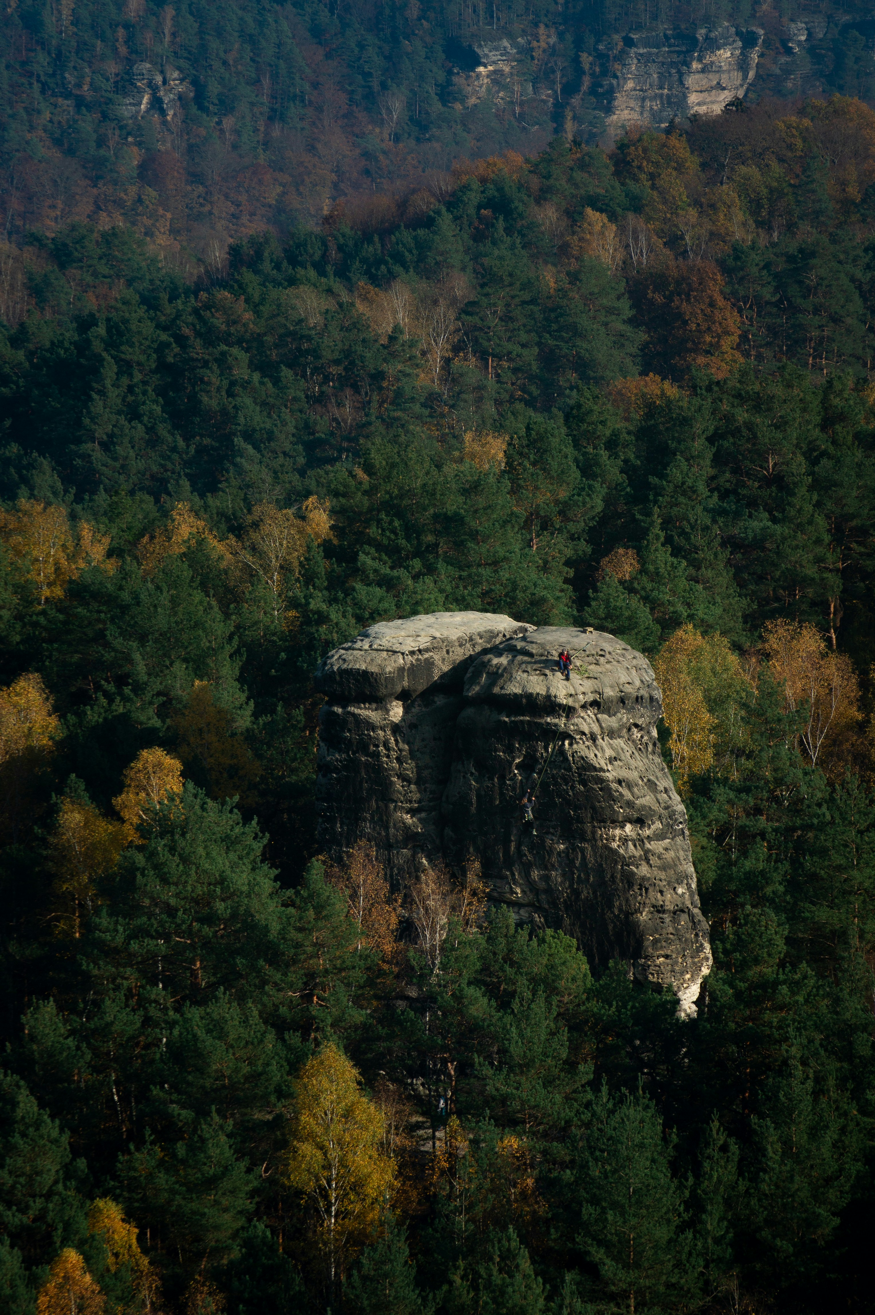 A towering rock formation rises amidst a sea of autumn foliage, with a climber scaling its rugged surface. The scene captures the harmony between nature and adventure.