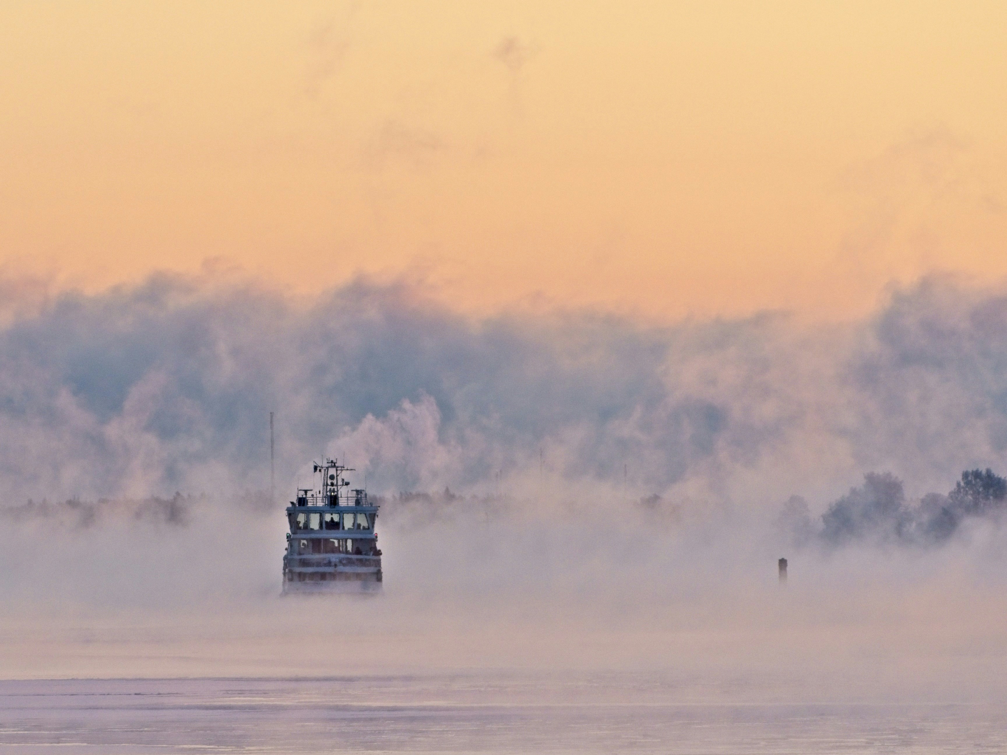 A boat navigates through thick fog on a calm river at dawn, surrounded by ethereal mist and soft pastel hues in the sky.