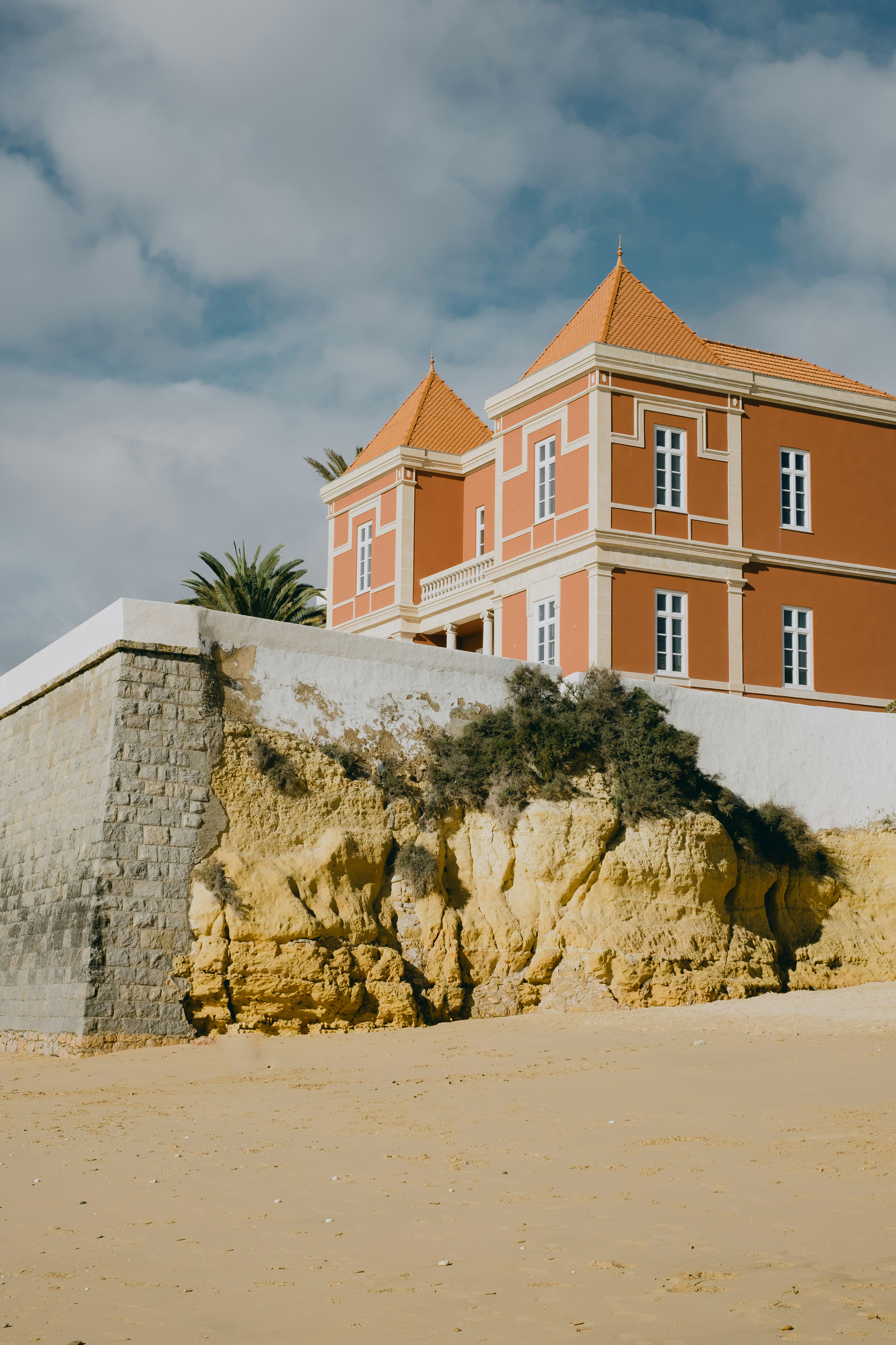 Historic coastal building with orange façade and pointed rooftops, perched above a sandy beach and rocky cliffs.
