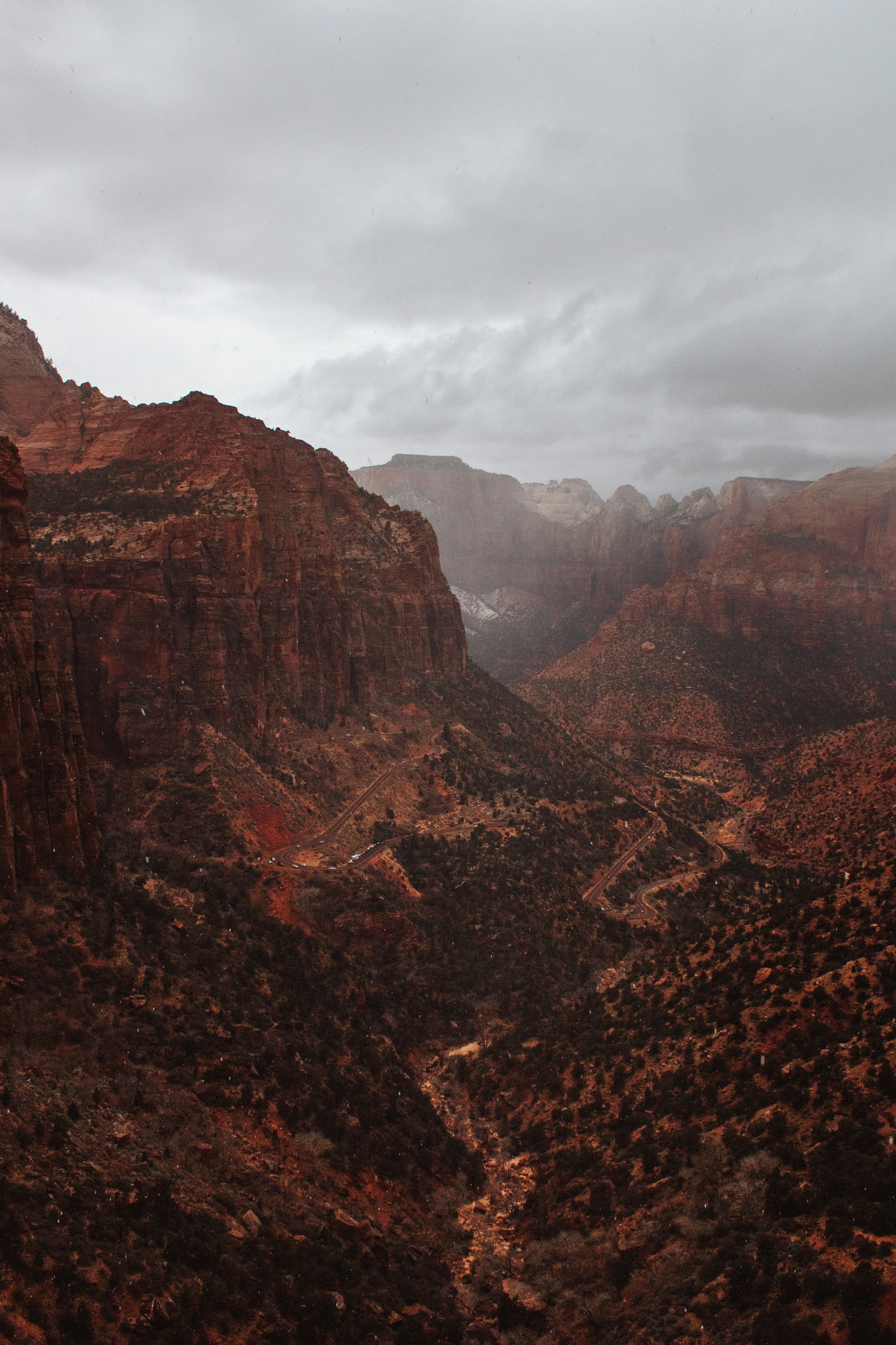 Majestic red rock formations loom over a winding valley, shrouded in mist under a cloudy sky.