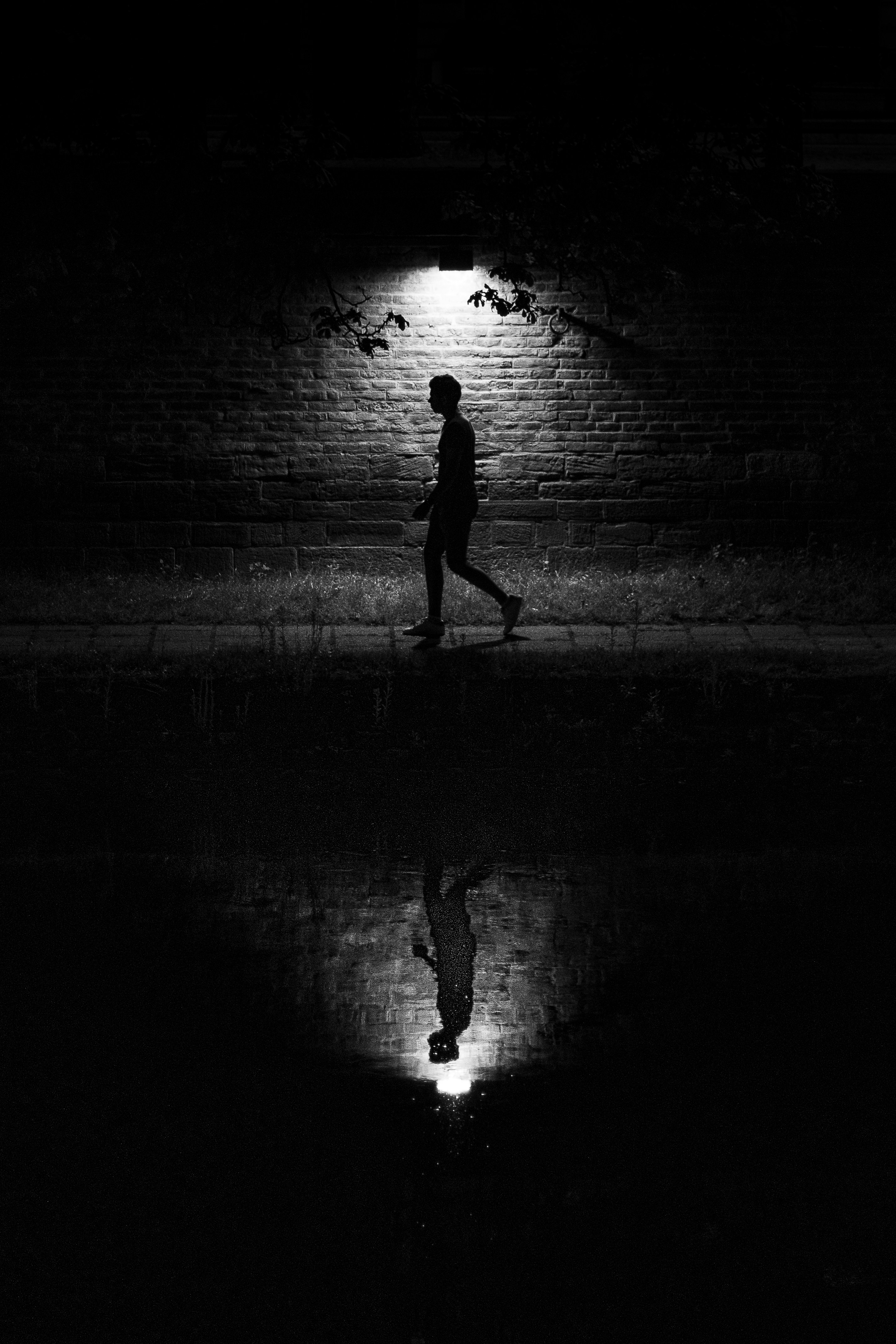 Silhouette of a person walking under a streetlight with their reflection mirrored on a canal at night.
