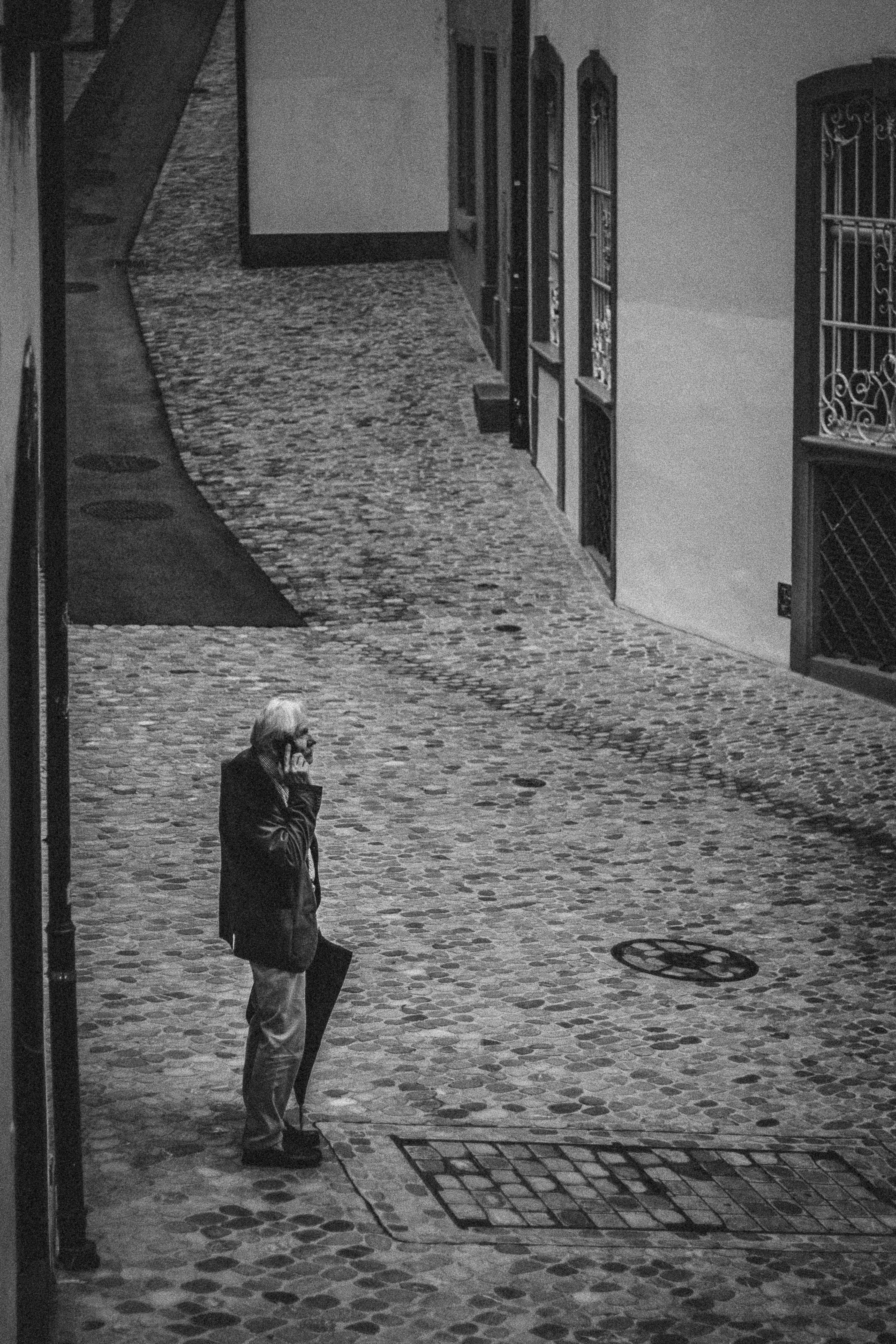 An elderly man stands alone on a cobblestone street, holding an umbrella and deep in thought. The narrow alleyways create a sense of isolation.