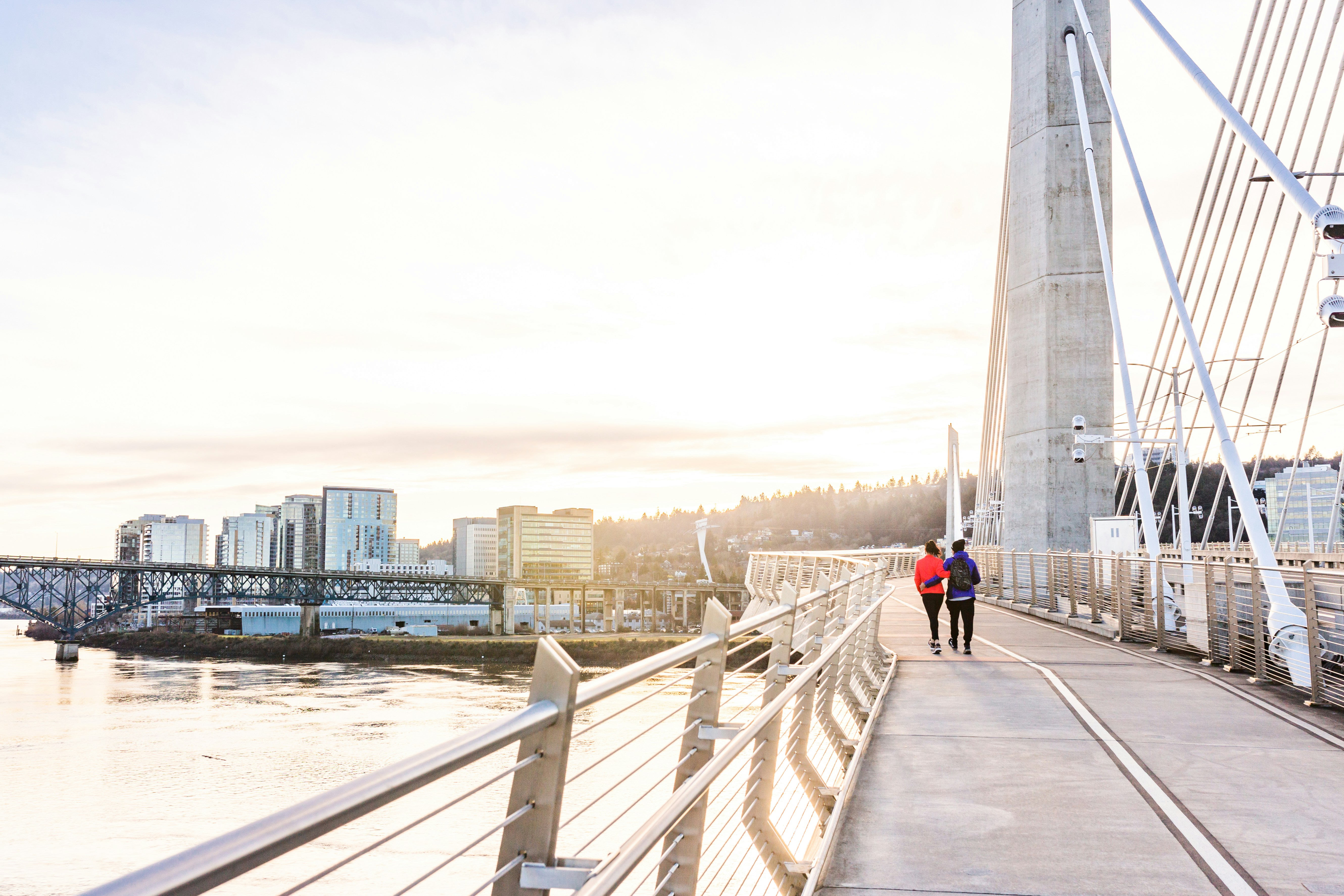 Two individuals walking on a modern pedestrian bridge with city buildings in the background during sunset.