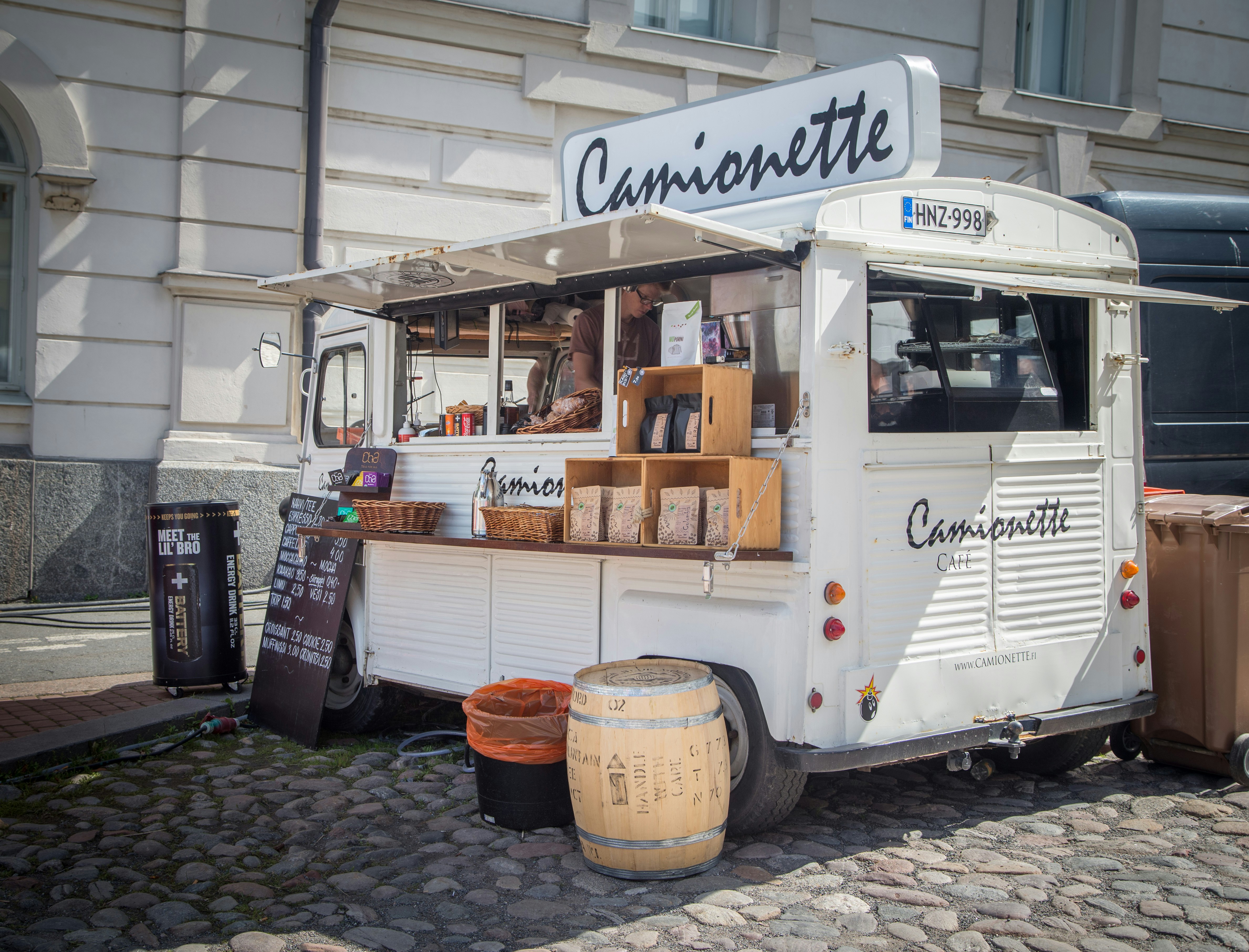 Vintage coffee van serving drinks on a cobblestone street with wooden barrels and boxes.