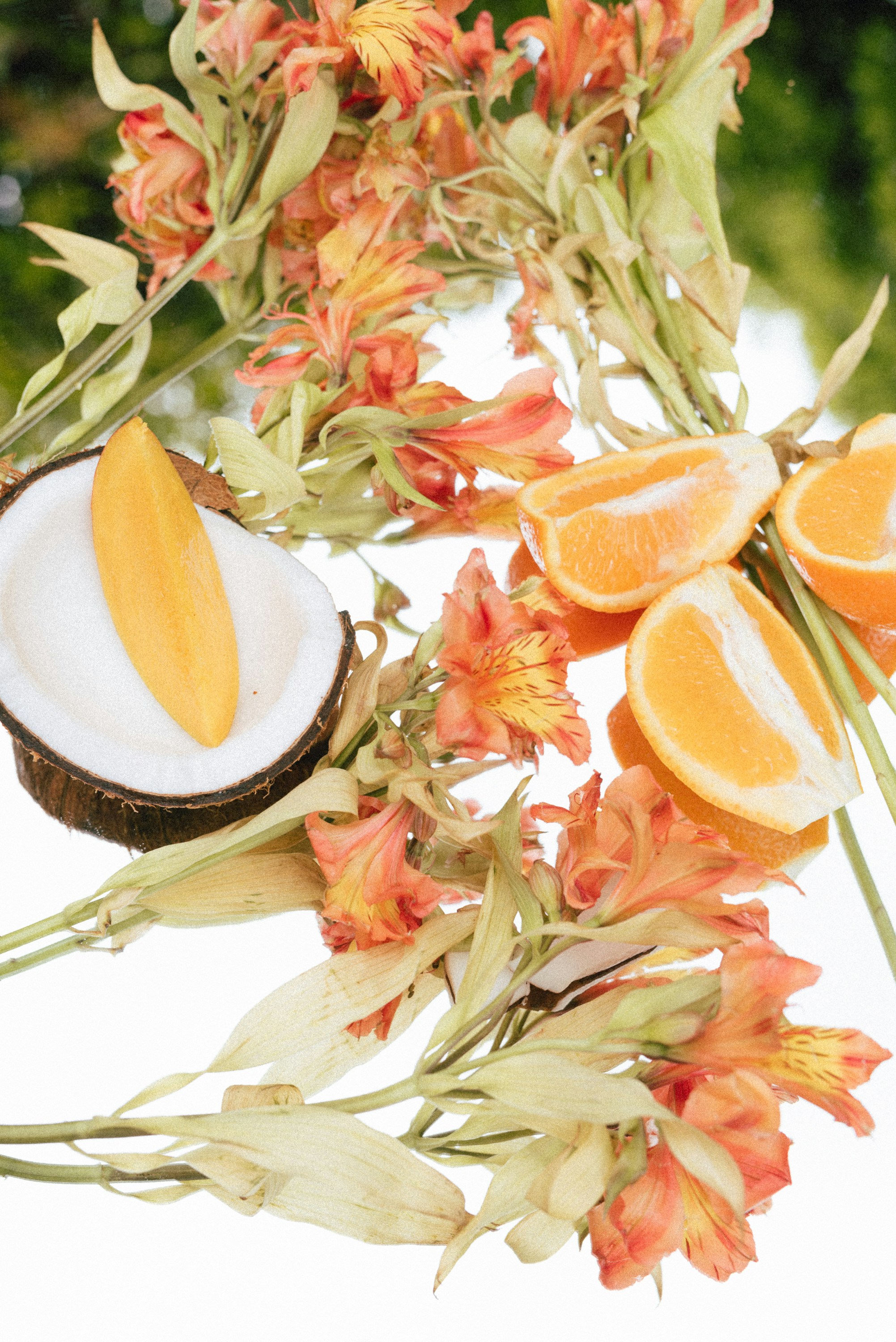 Arrangement of vibrant flowers, coconut, and orange segments artfully displayed on a reflective surface.