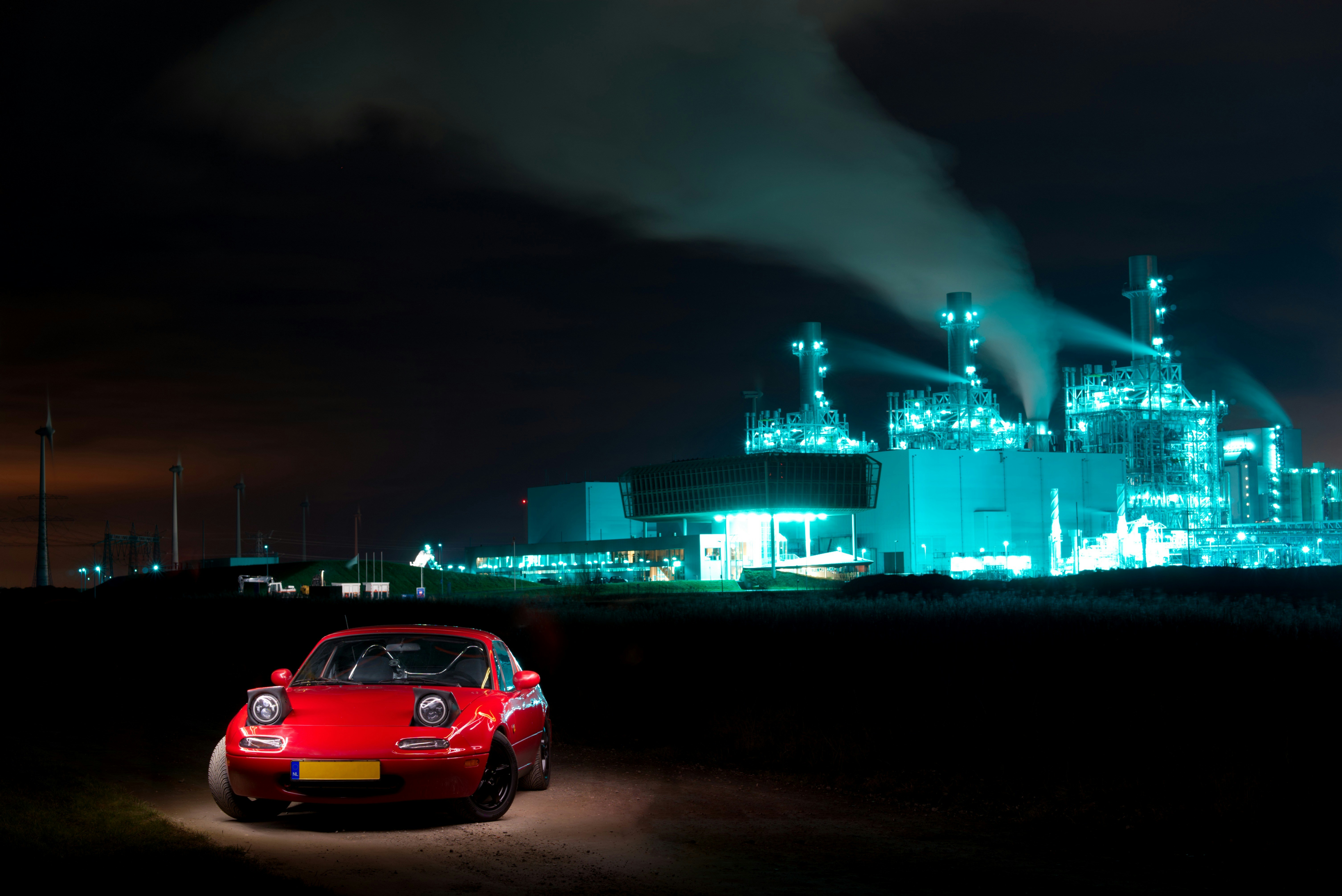 A vintage red sports car parked on a dirt road with a glowing industrial complex in the background, illuminated by vibrant blue lights and smoke trails.
