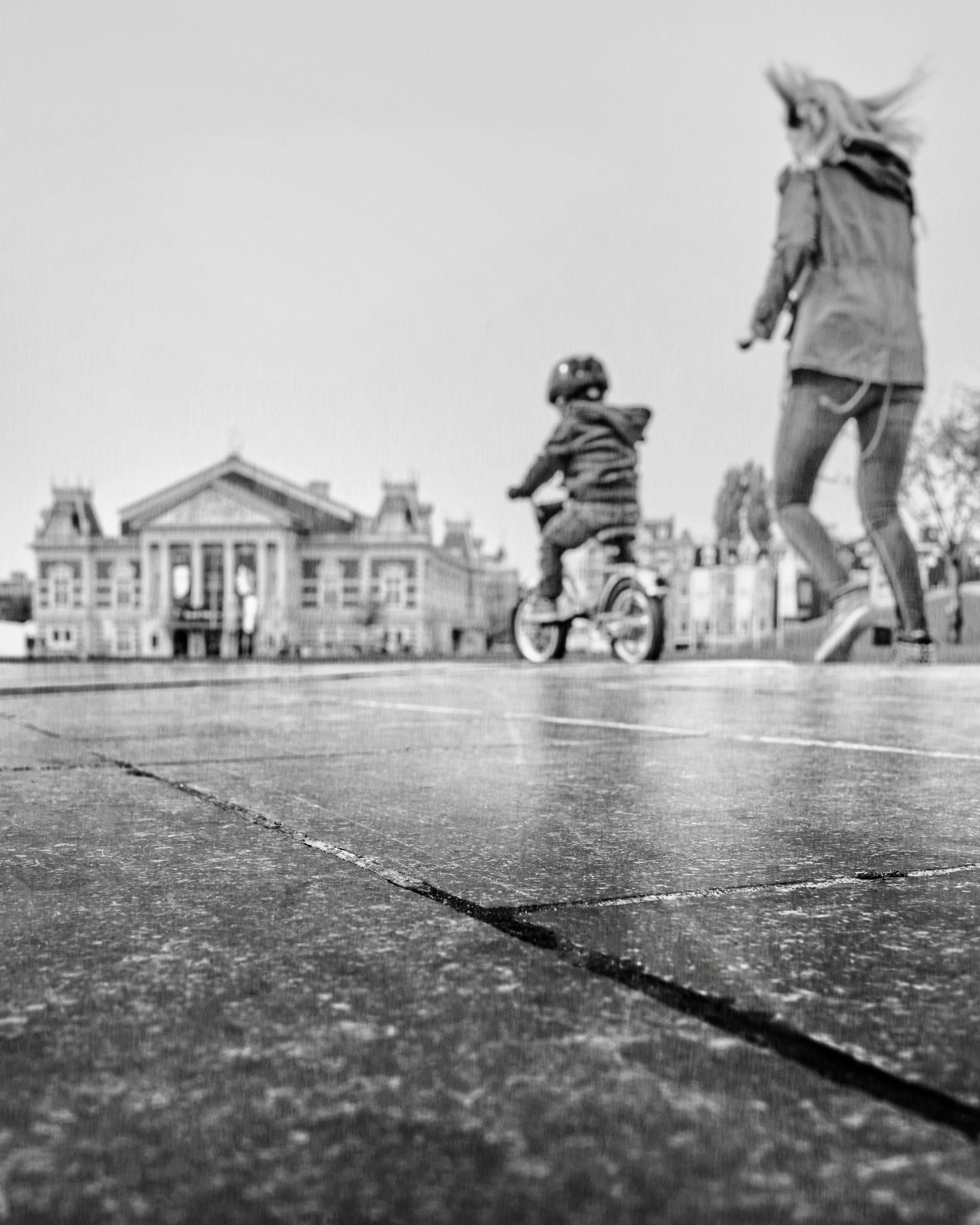 Child riding a balance bike in a city square, with a woman walking nearby and historic architecture in the background.