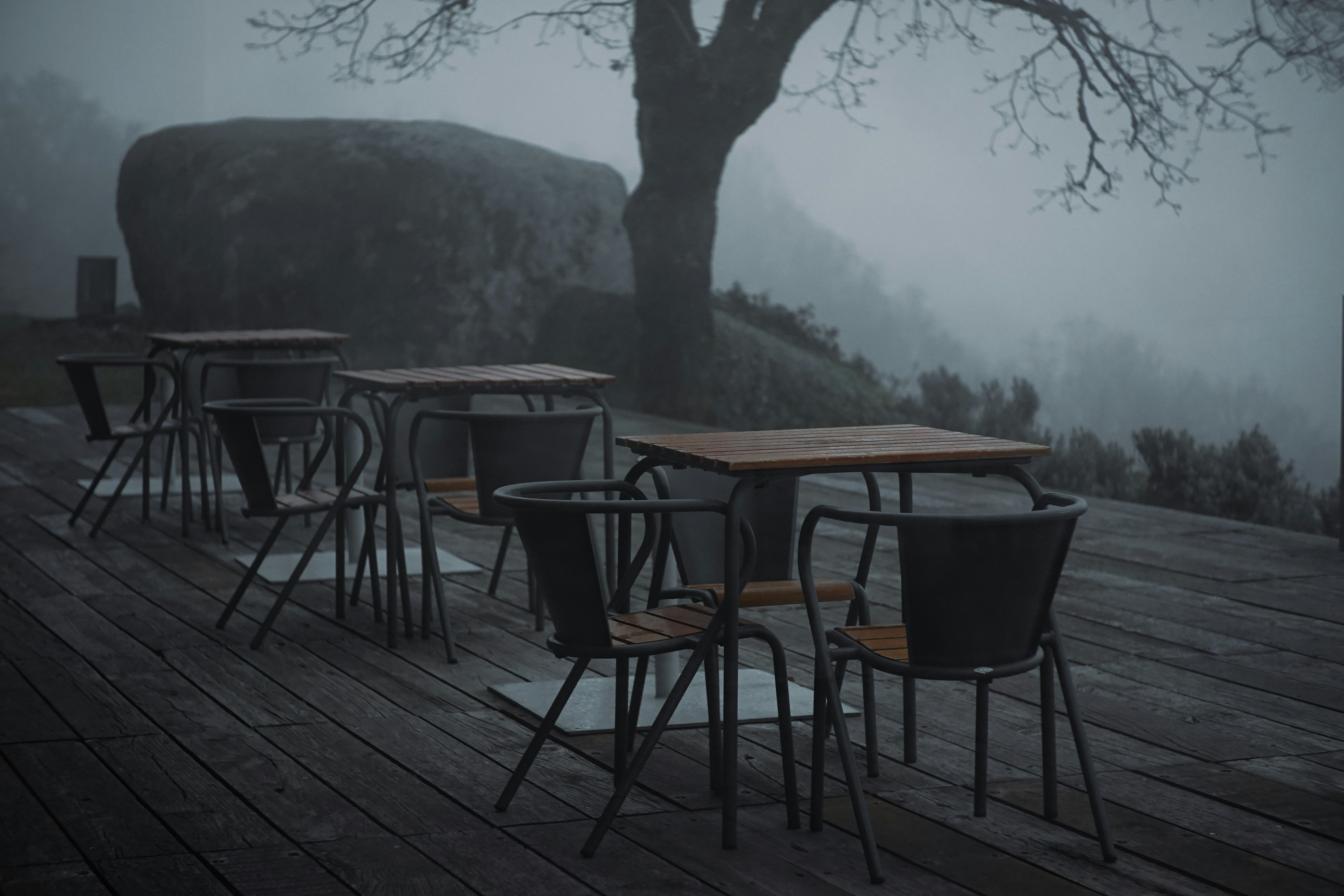 Empty tables and chairs on a wooden deck surrounded by misty landscapes, evoking a sense of tranquility and isolation. The scene captures the essence of a quiet getaway.