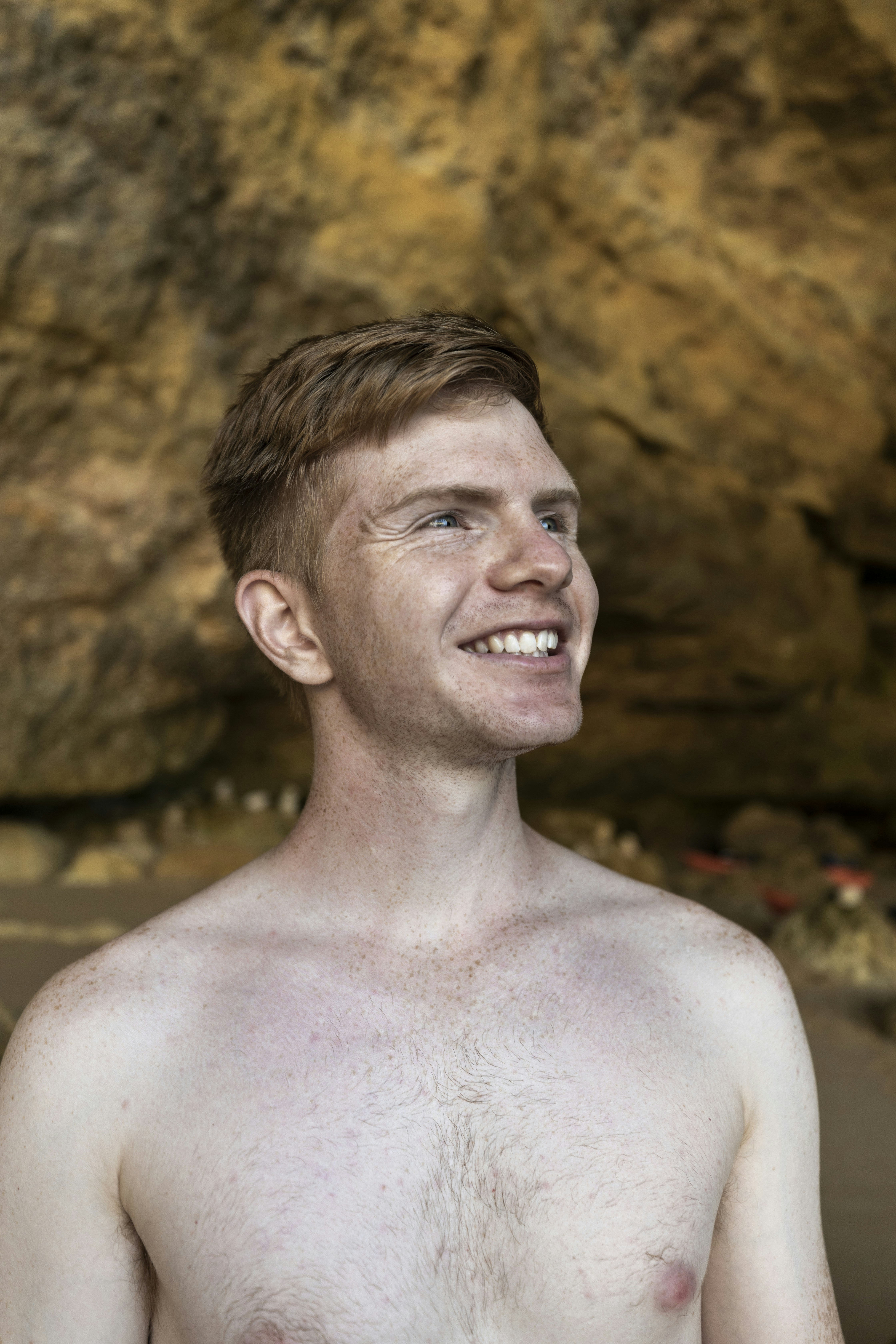 A smiling young man stands under a rocky overhang, exuding happiness and warmth. His relaxed demeanor complements the natural beauty surrounding him.